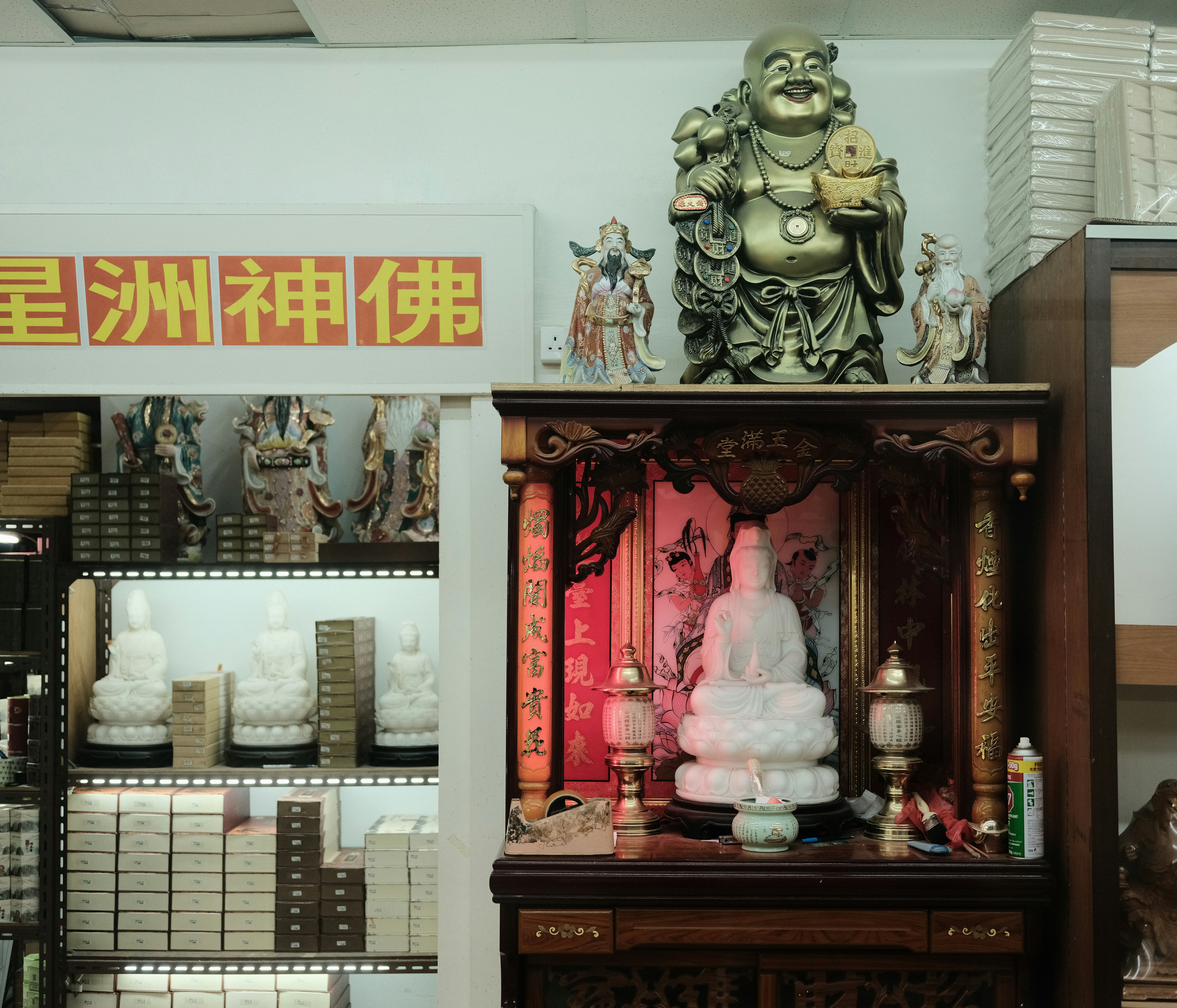 A tranquil altar featuring a white Buddha statue surrounded by intricate carvings and a smiling figure above, set against a backdrop of decorative elements and shelves filled with artifacts.