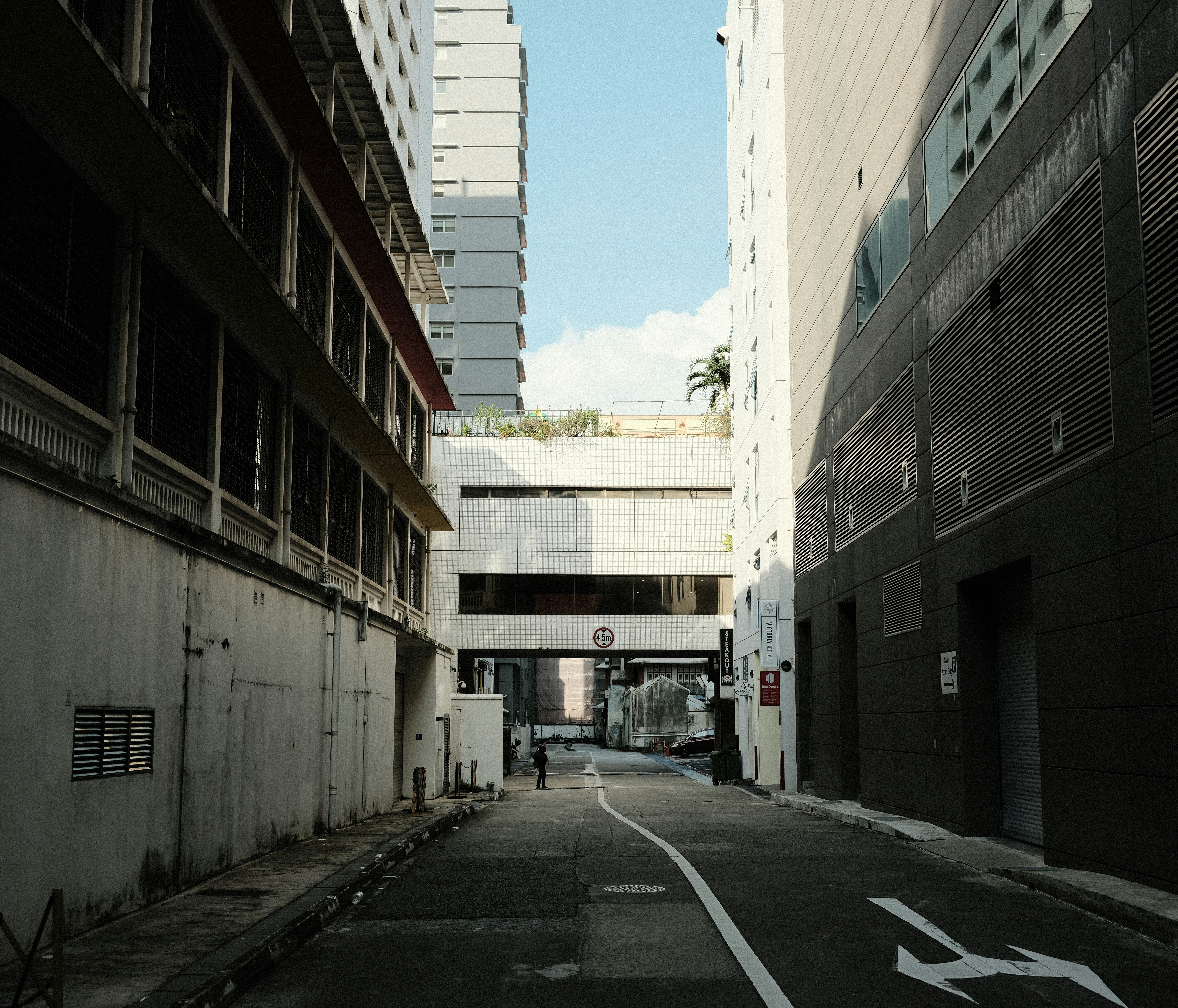 Narrow alleyway flanked by modern buildings, leading towards a bright sky. The scene captures the essence of urban architecture and the interplay of light and shadow.