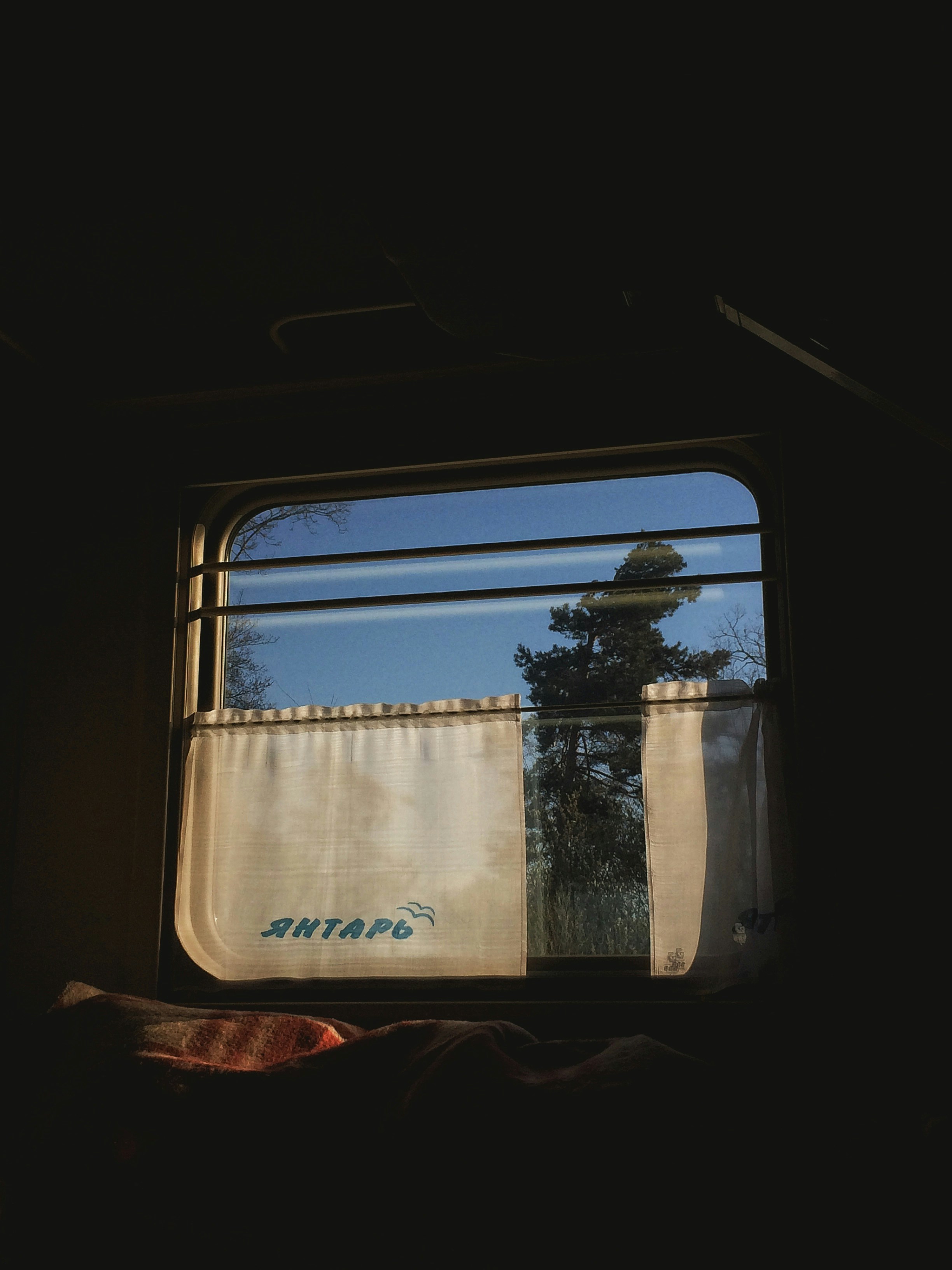 Sunlight streaming through a train window, illuminating sheer curtains and revealing a serene outdoor scene with trees against a clear blue sky.