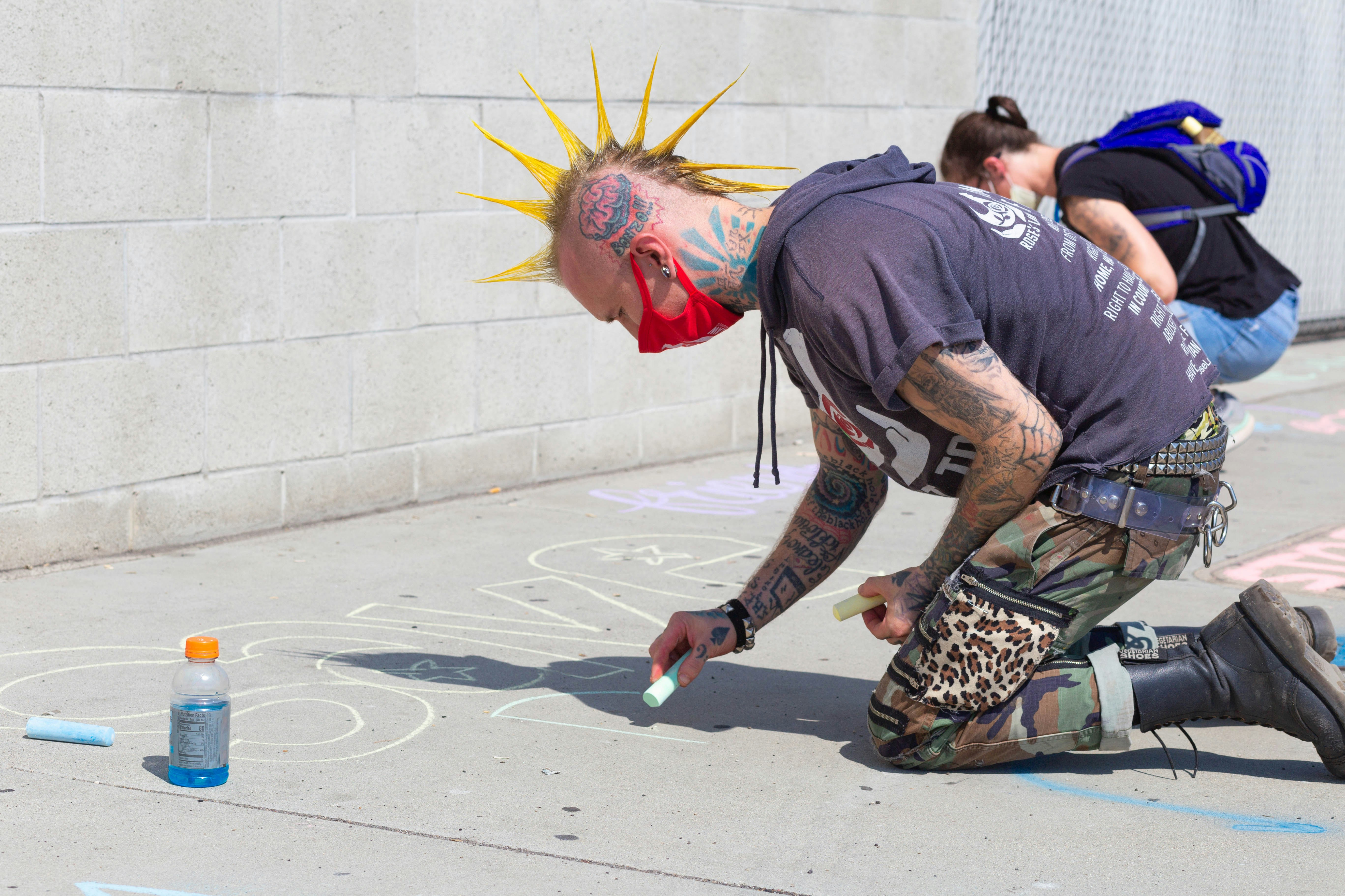 Tattooed individual with spiked yellow hair creating chalk art on a concrete sidewalk.