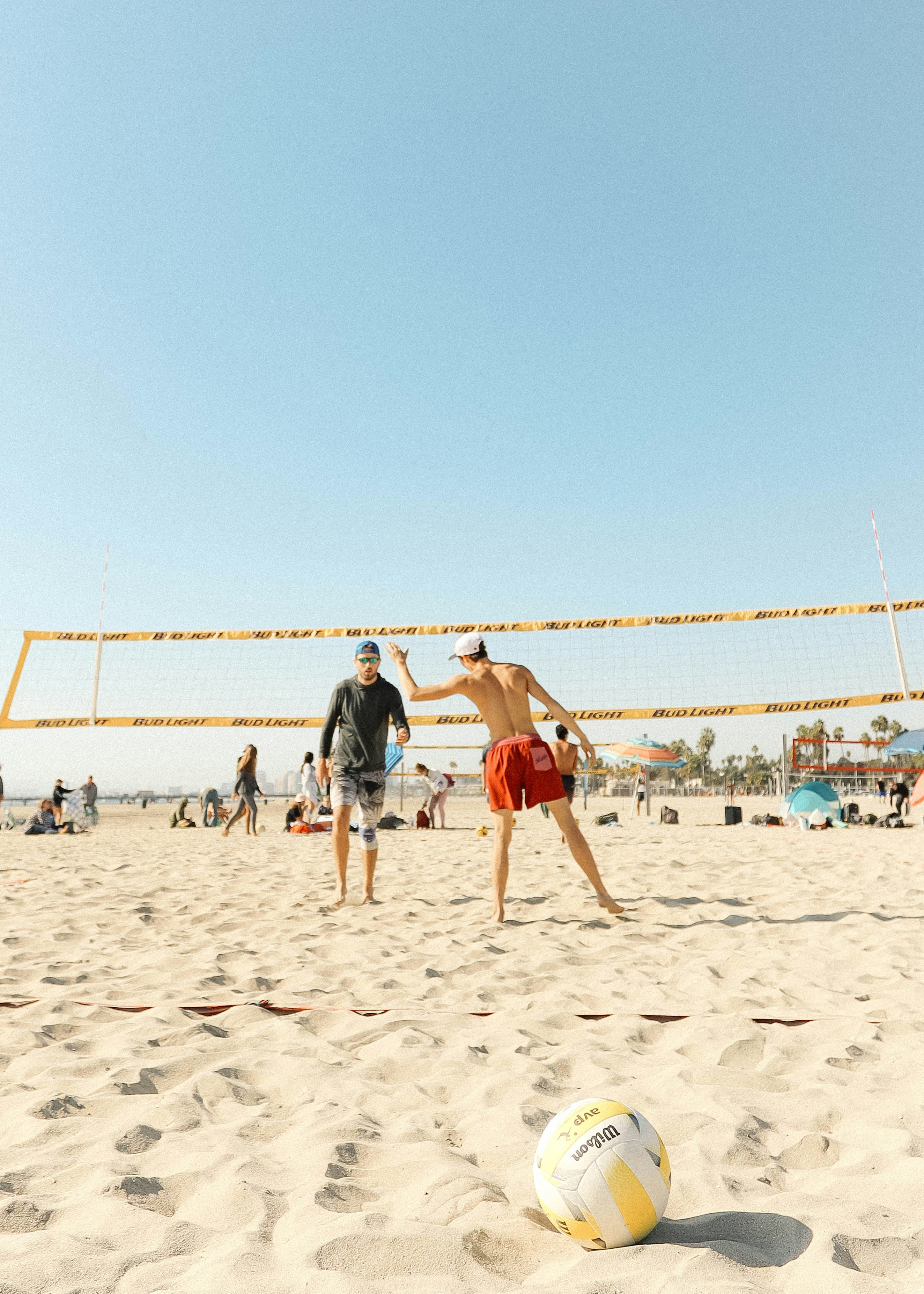 Two players engaging in a lively beach volleyball match, with a ball resting on the sand in the foreground. The vibrant beach atmosphere is evident with spectators in the background.