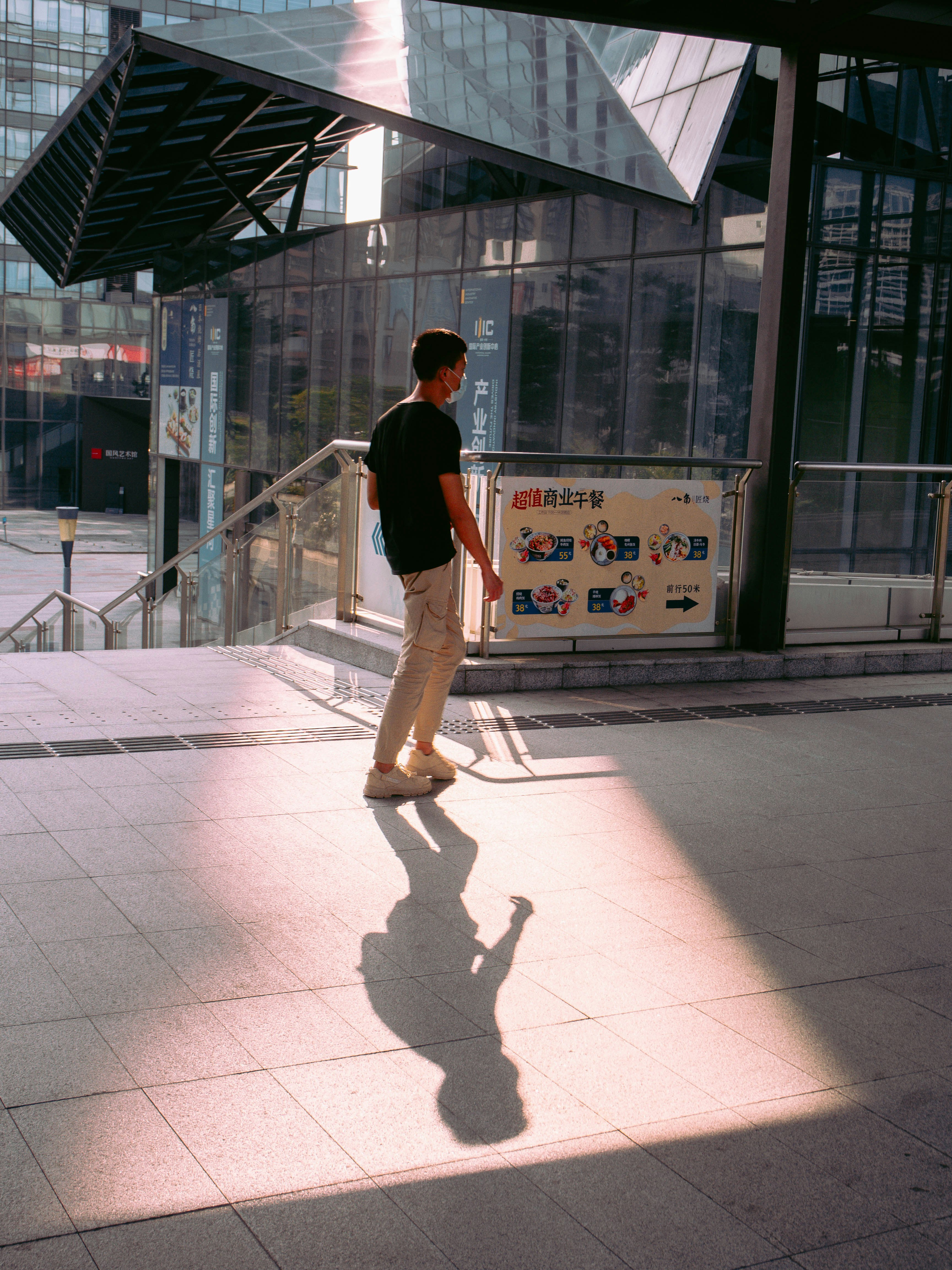 A figure walks through a sunlit urban space, casting a long shadow on the pavement. The scene captures the interplay of light and shadow against a modern architectural backdrop.
