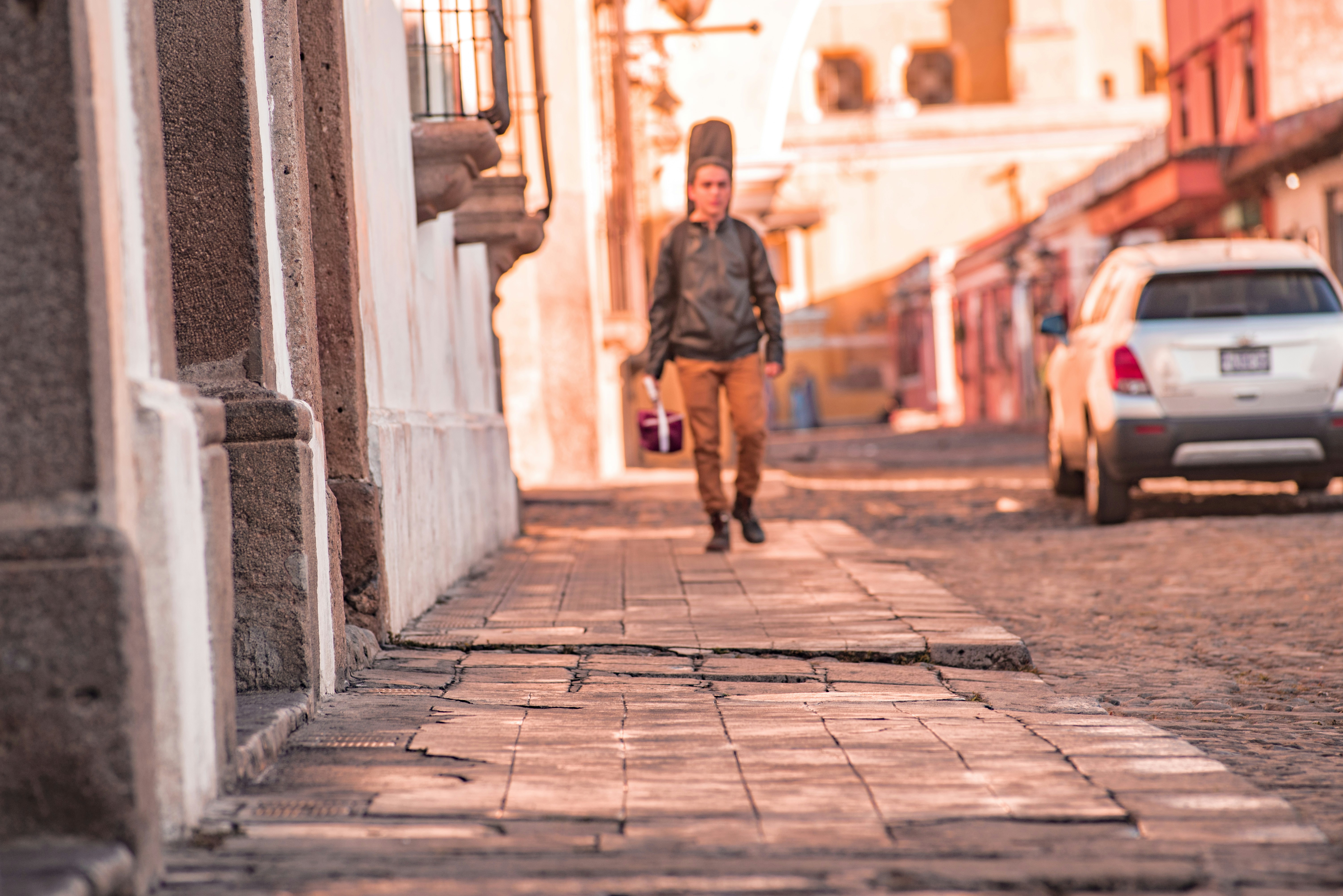 A person walks down a cobblestone street lined with colorful buildings, carrying a bag. The warm light of the setting sun casts a soft glow on the scene.