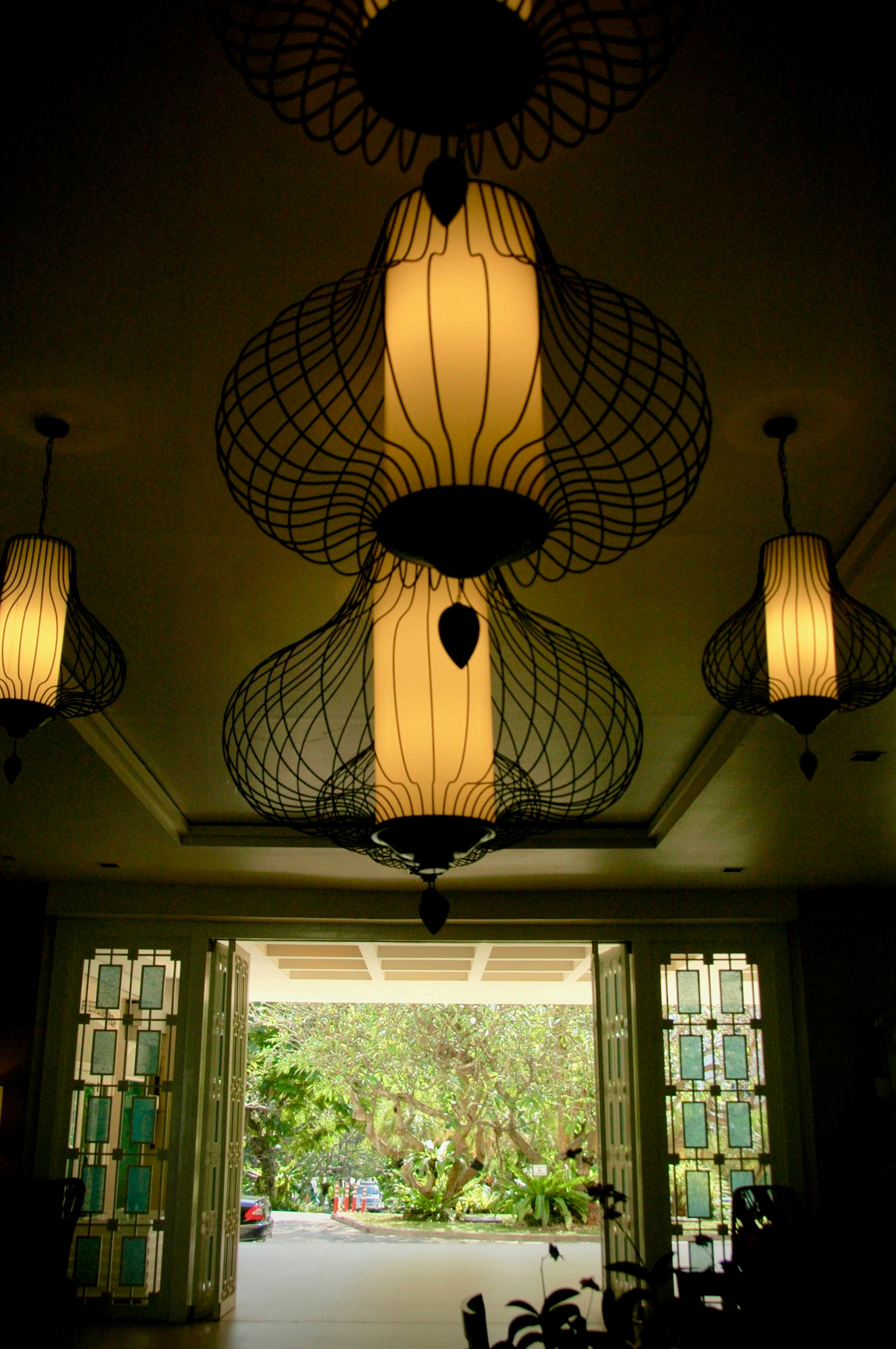 Intricately designed lanterns hang from the ceiling, framing an open doorway that leads to a lush garden. The interplay of light and shadow creates a serene atmosphere.