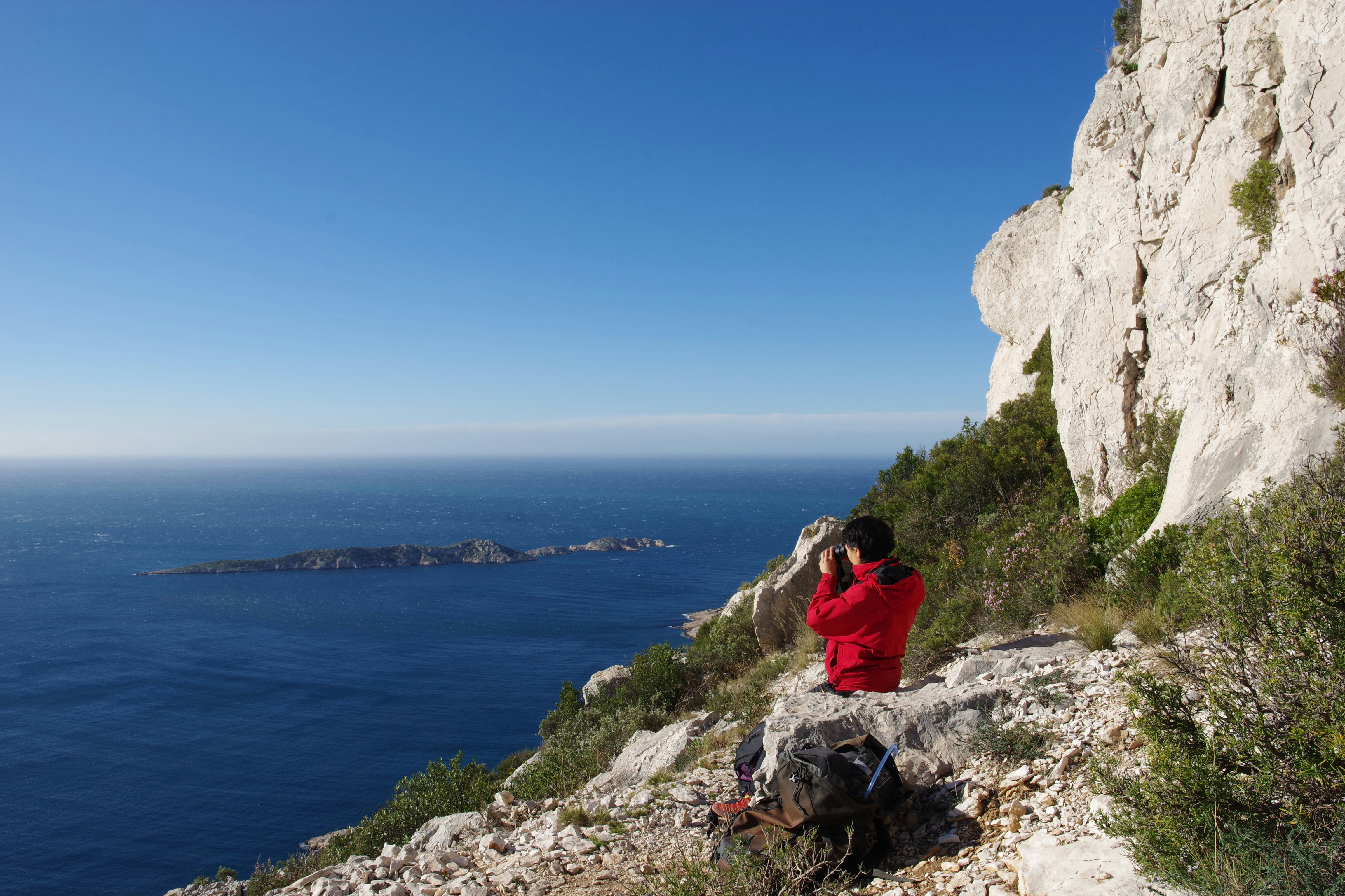 Crimson-clad Photographer on a Sunlit Cliff Overlooking the Azure Sea