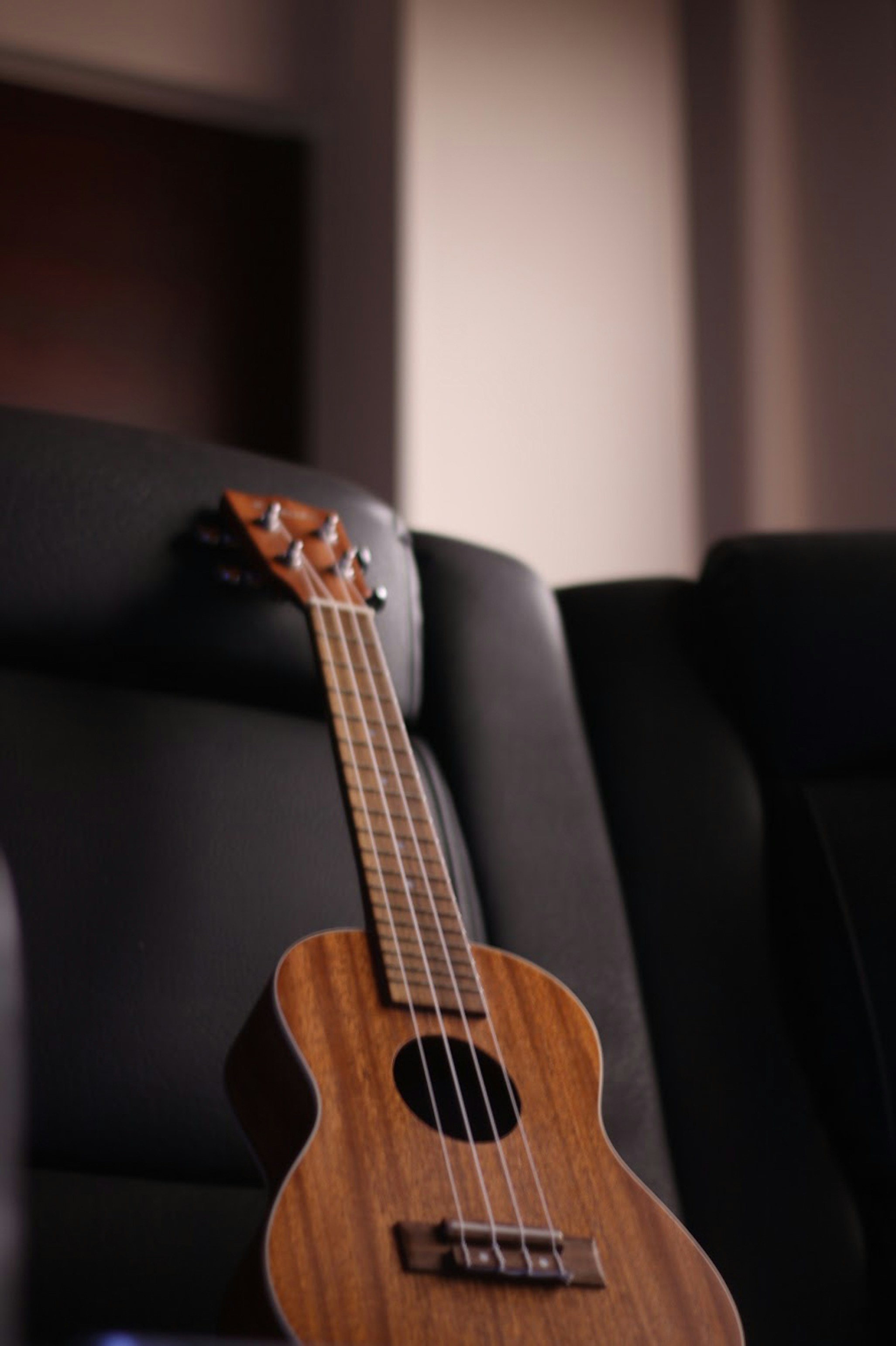 A wooden ukulele rests against a black leather chair, showcasing its smooth curves and detailed craftsmanship.
