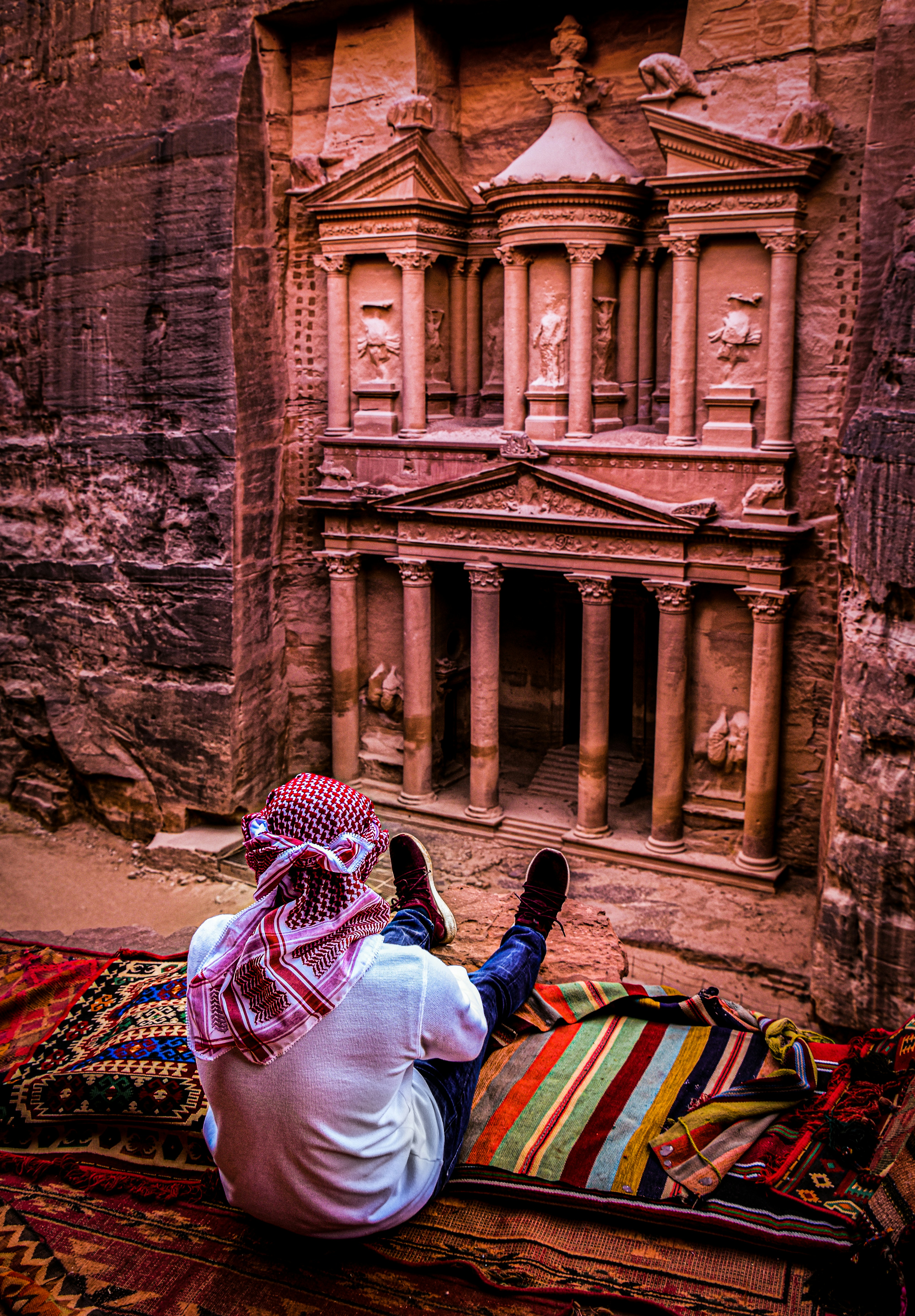 Person in traditional attire seated on colorful rugs, gazing at the rock-carved facade of a historical monument.