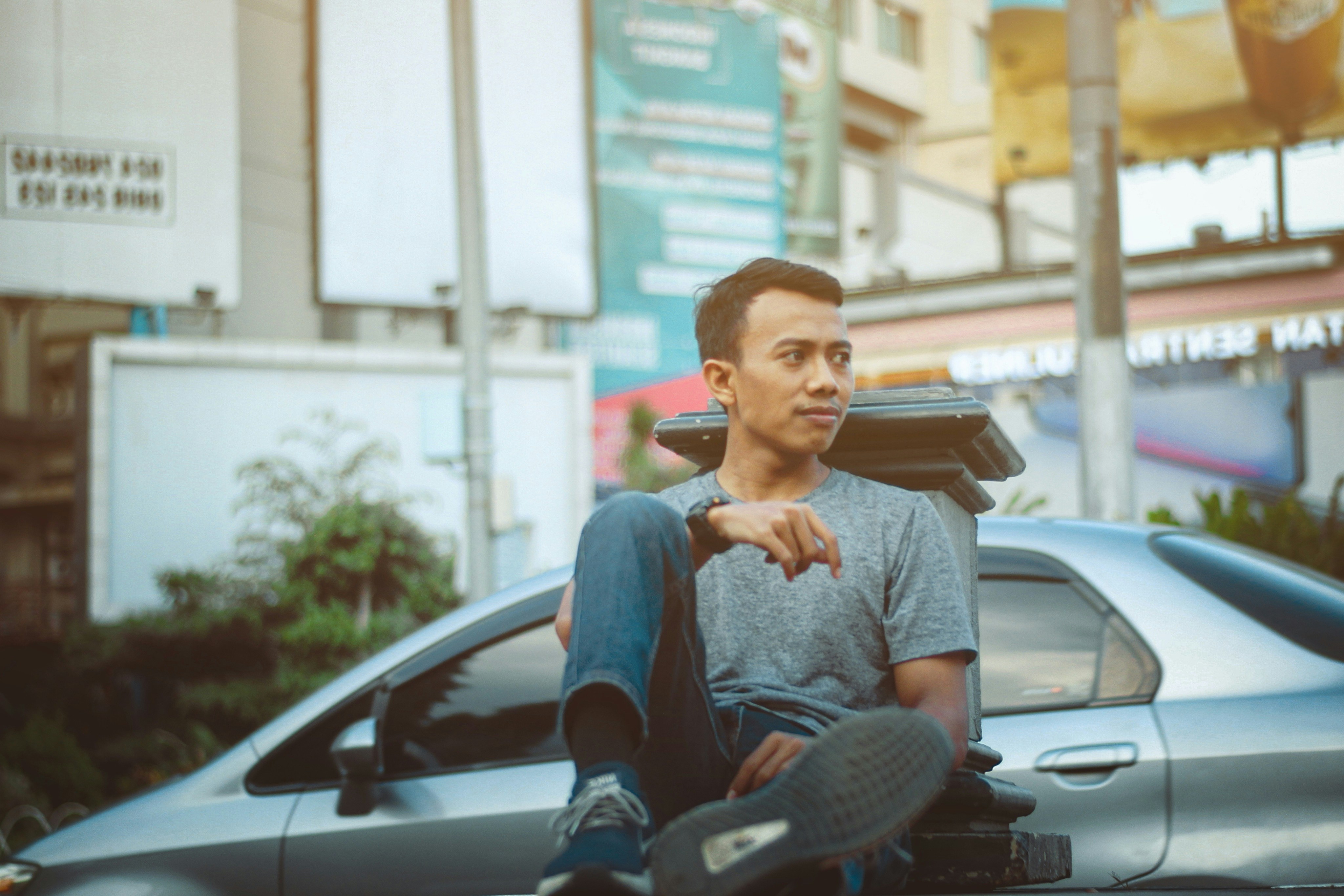 Young man casually seated on a car in an urban setting, surrounded by city life and greenery.