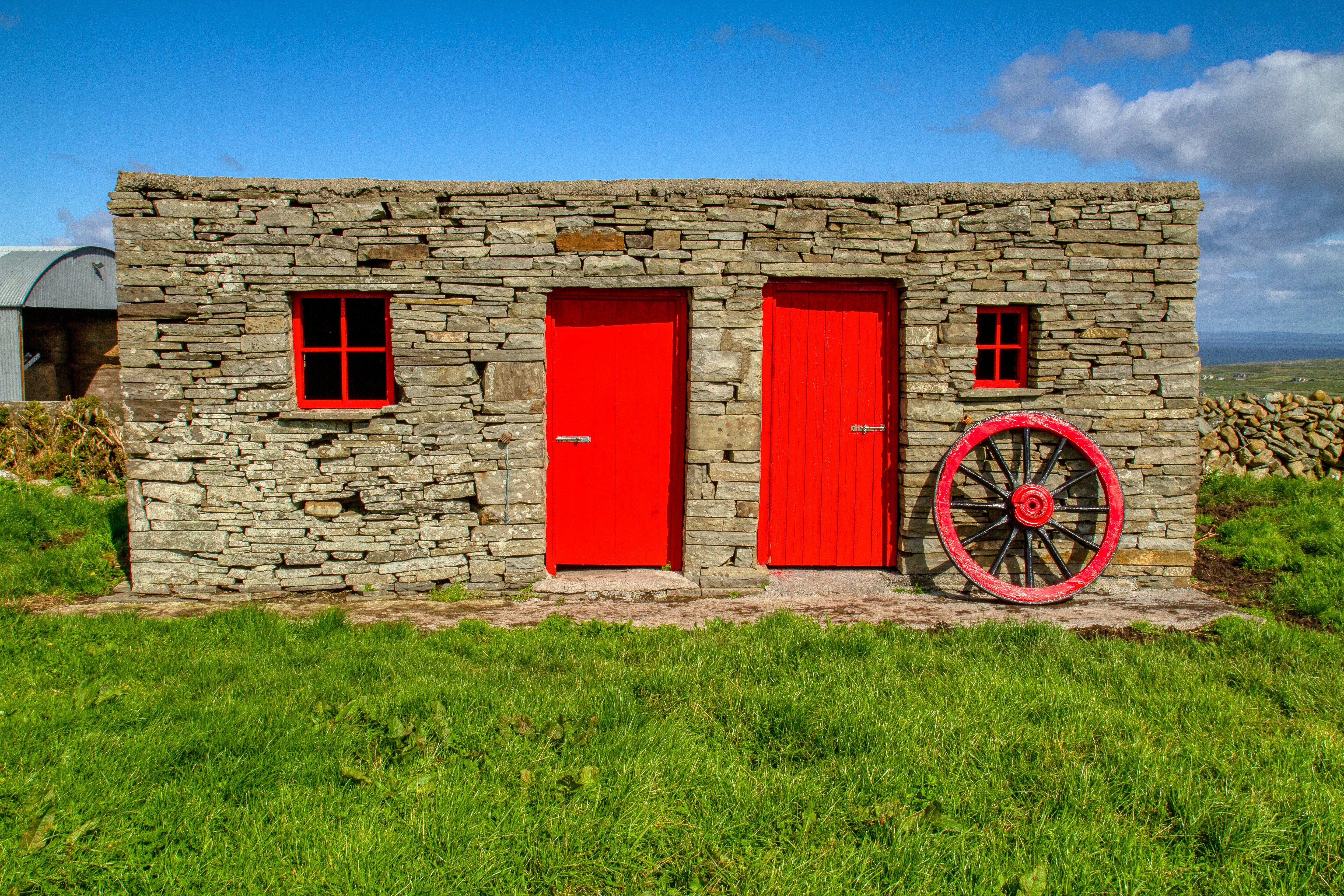 A weathered stone building featuring bright red doors and windows, accompanied by a decorative wheel, set against a lush green landscape.