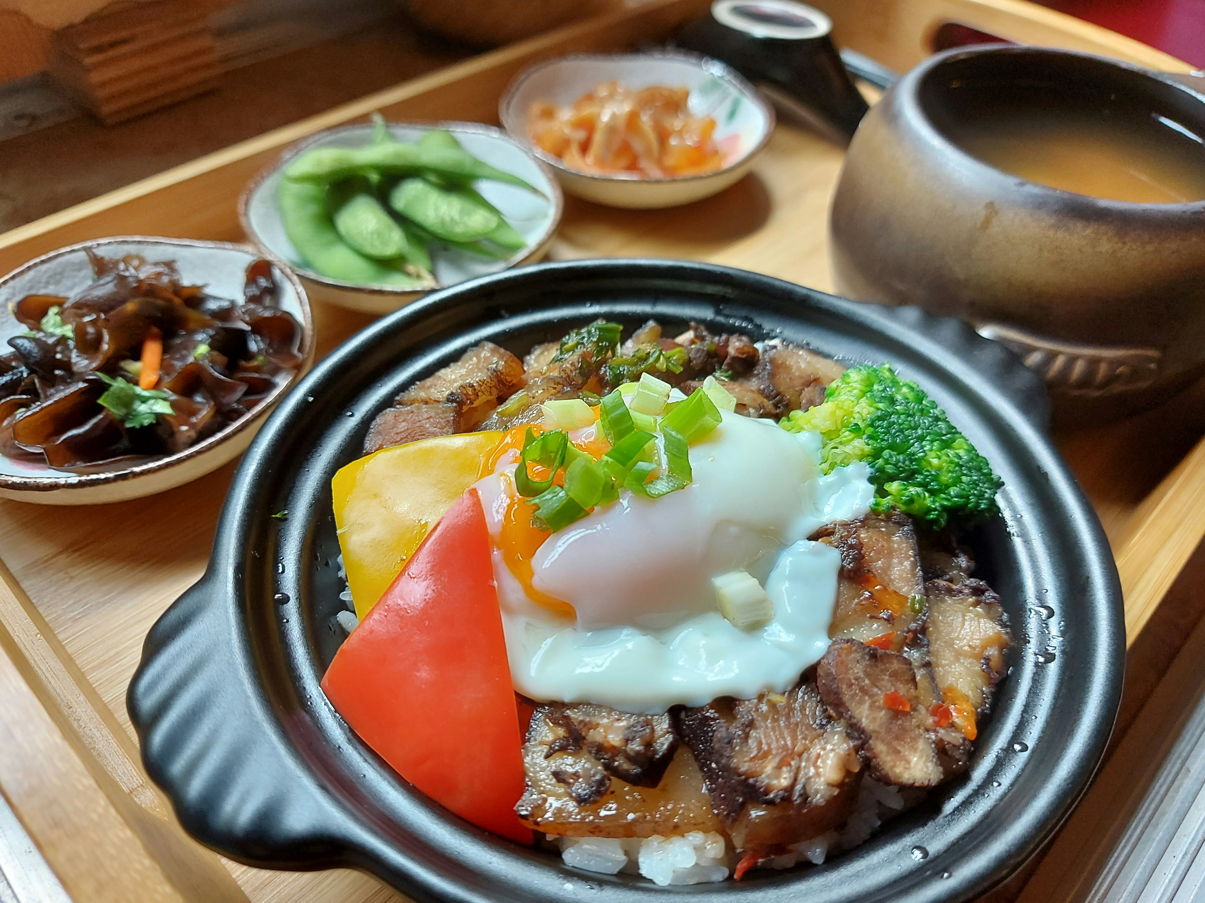 Vibrant rice bowl topped with grilled meat, vegetables, and a soft egg, accompanied by side dishes of pickled vegetables and soup.