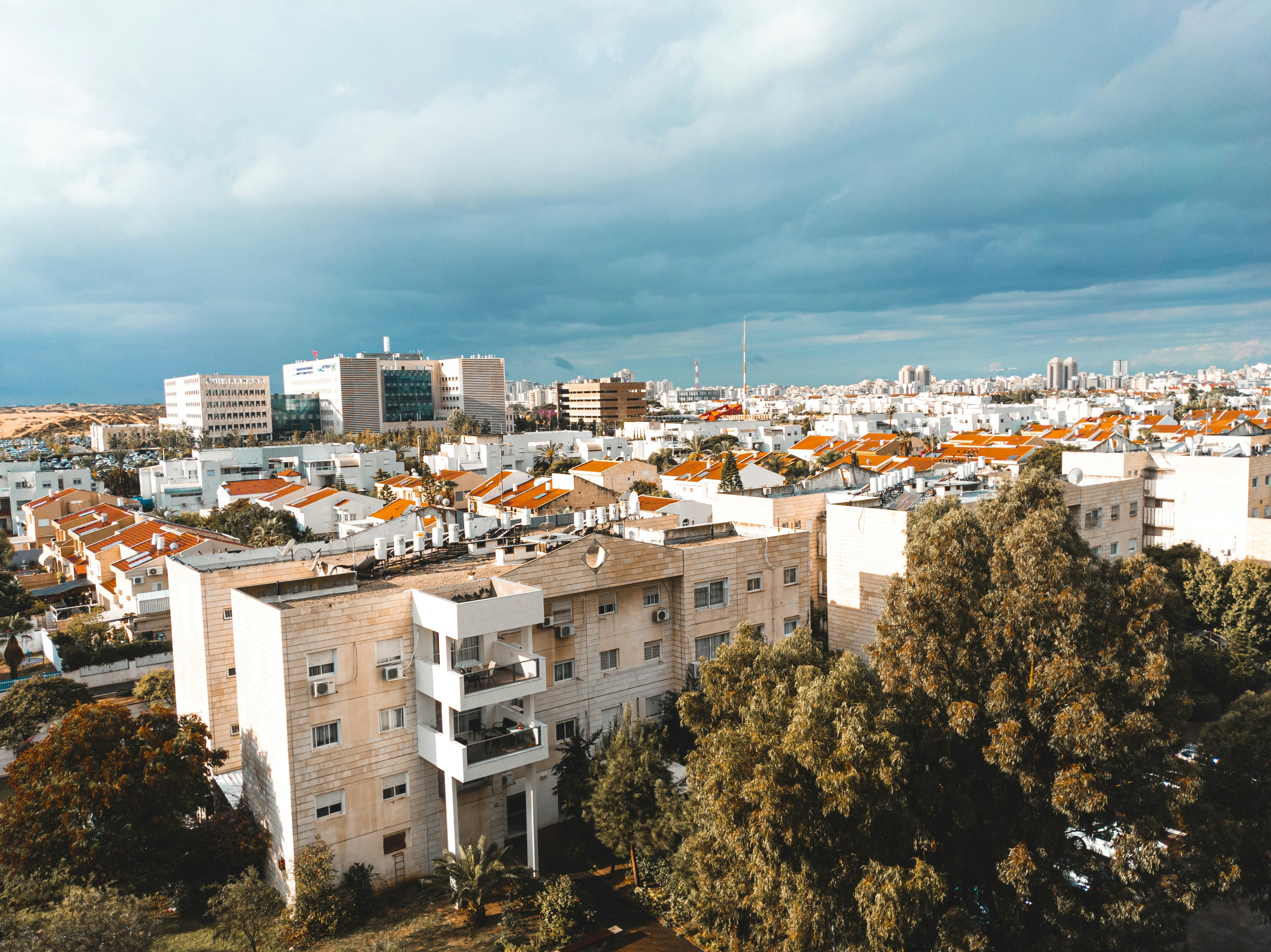 White concrete building surrounded by lush green trees with a sprawling cityscape in the background under a cloudy sky.