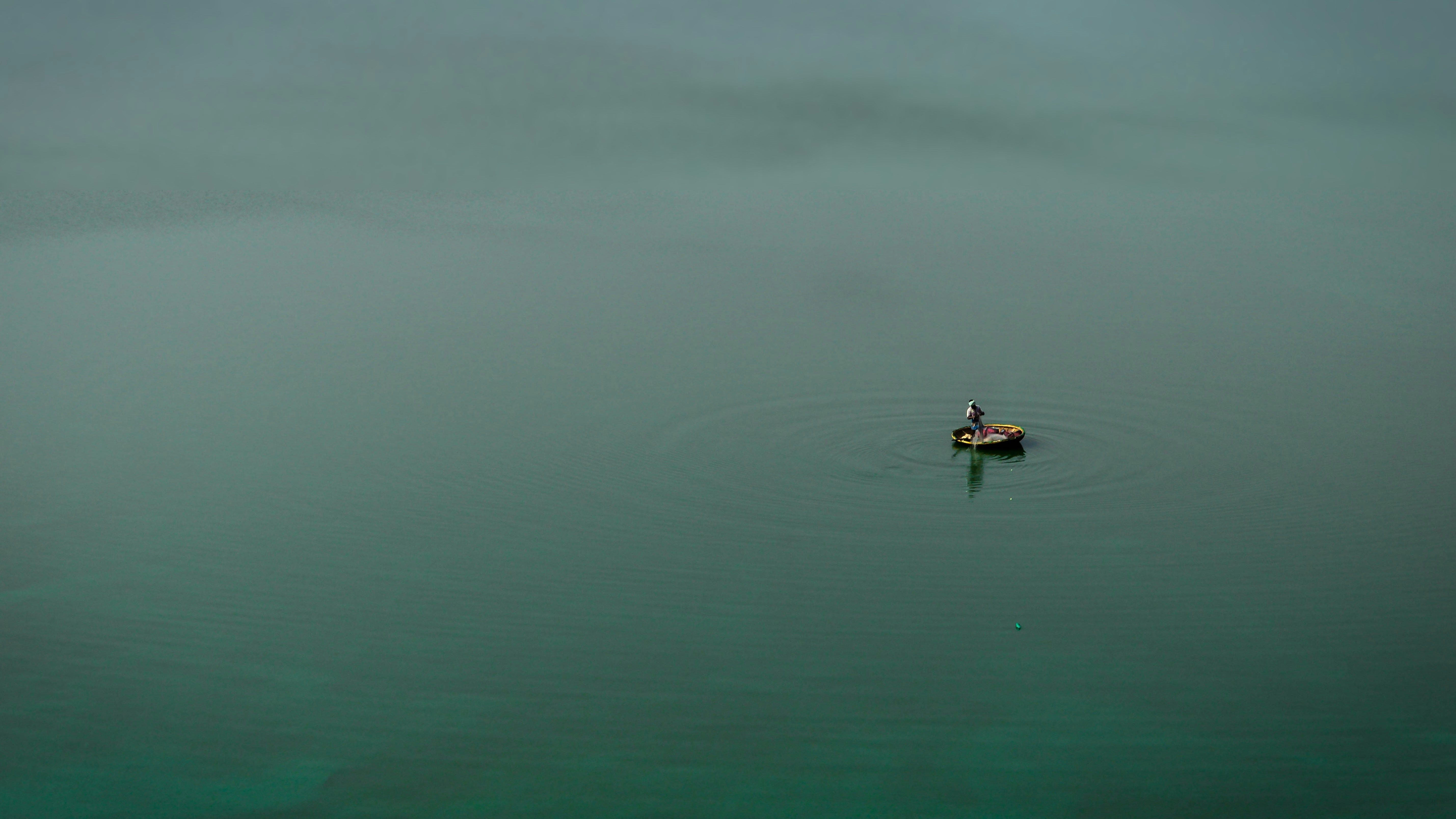 A lone figure in a small boat creates ripples on a tranquil body of water, surrounded by a misty atmosphere.
