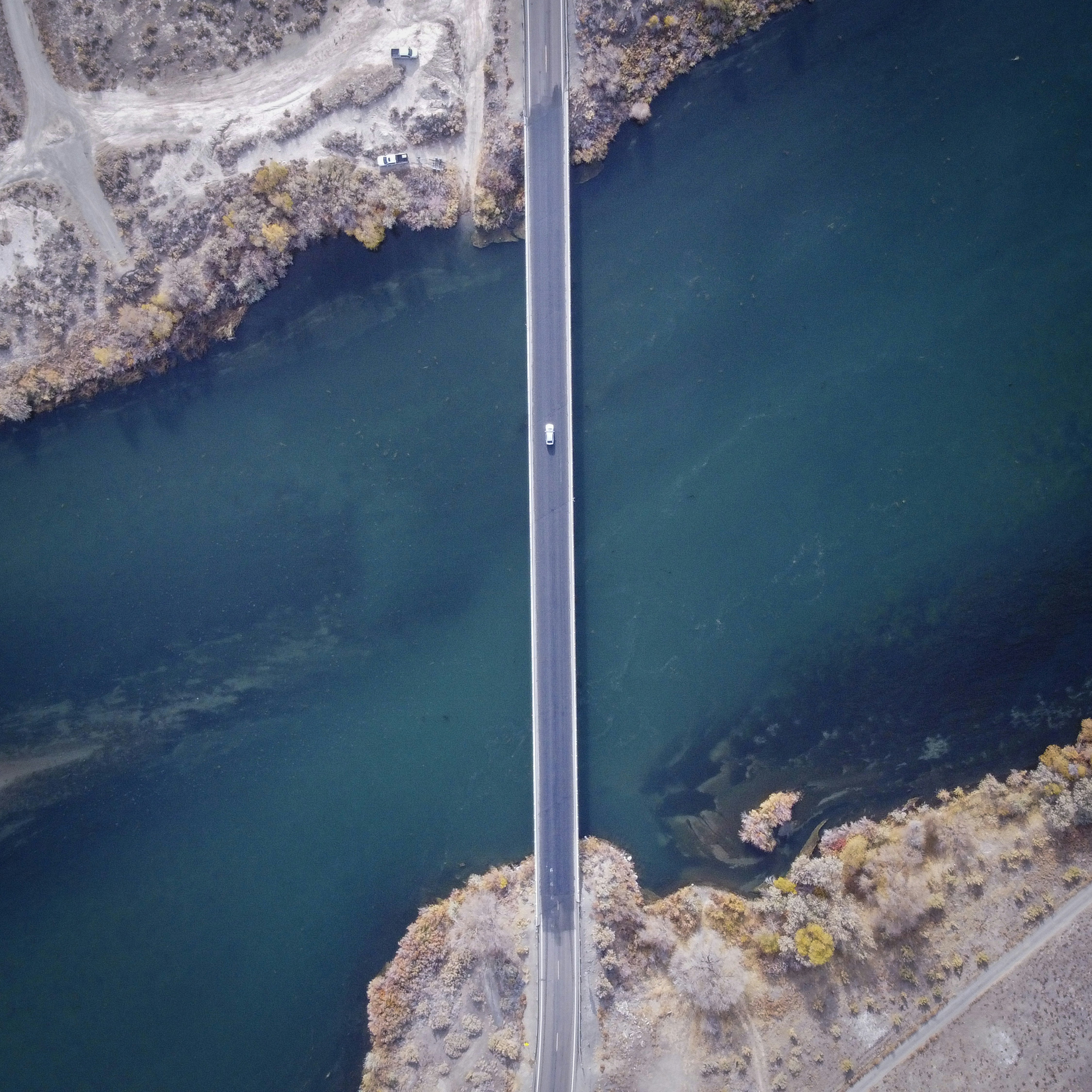 Concrete Spine Over Turquoise Currents: Aerial View of a Lone Car on a Riverside Bridge