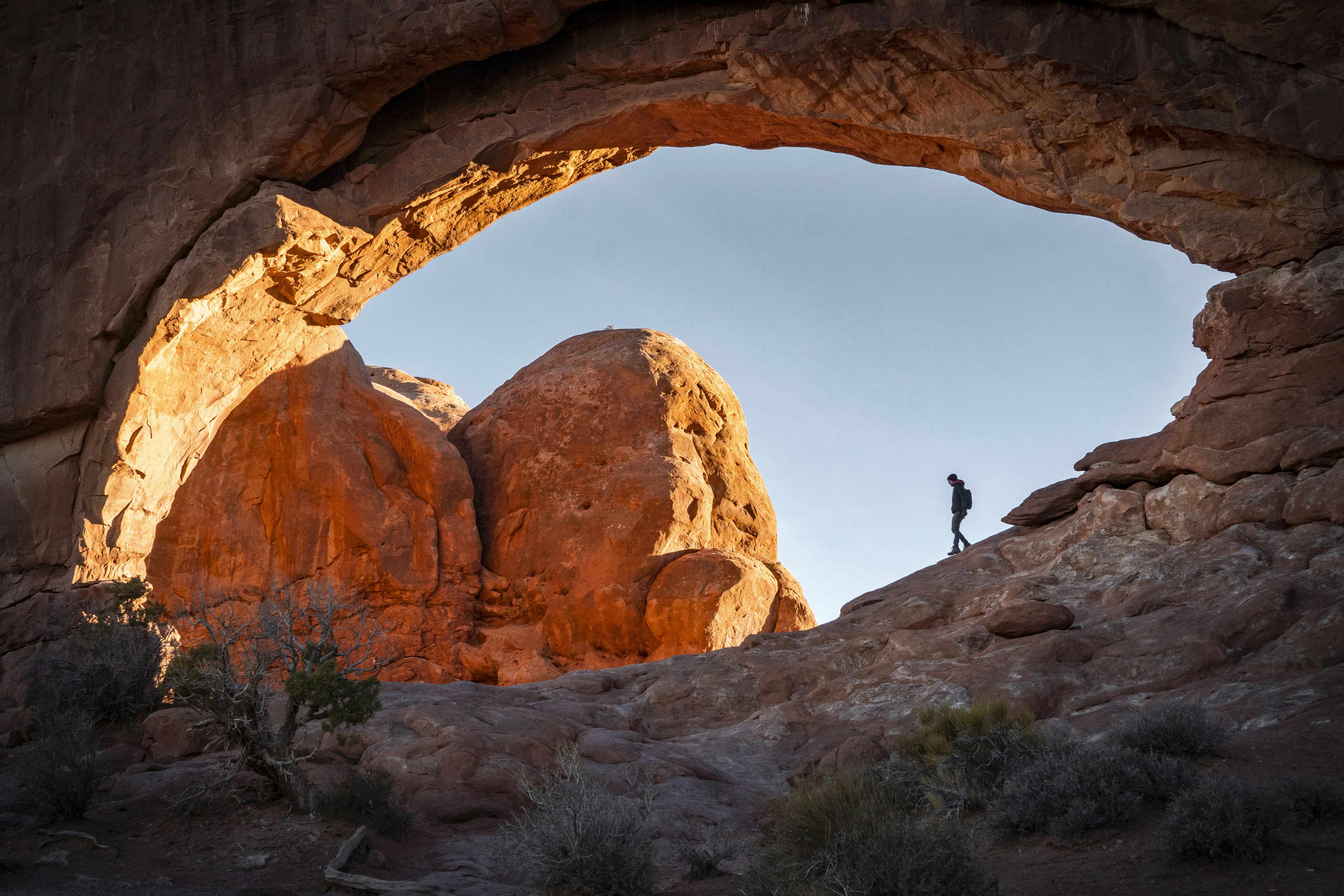 Hiker silhouetted against a natural arch, with warm sunlight illuminating the surrounding rock formations. The scene captures the essence of exploration in a rugged landscape.