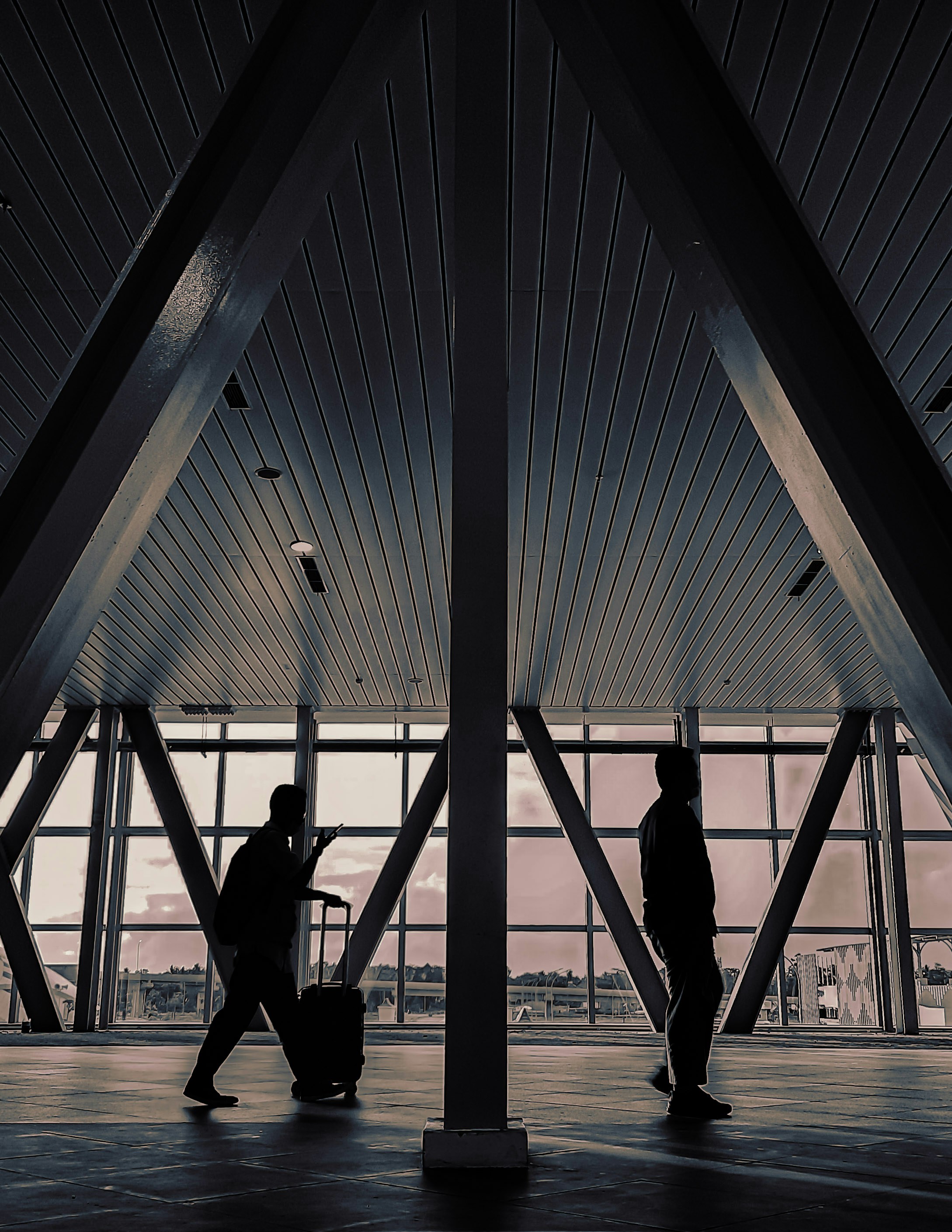 Low-light interior photograph of a terminal with diagonal steel beams forming an X pattern, two travelers silhouetted near expansive windows.