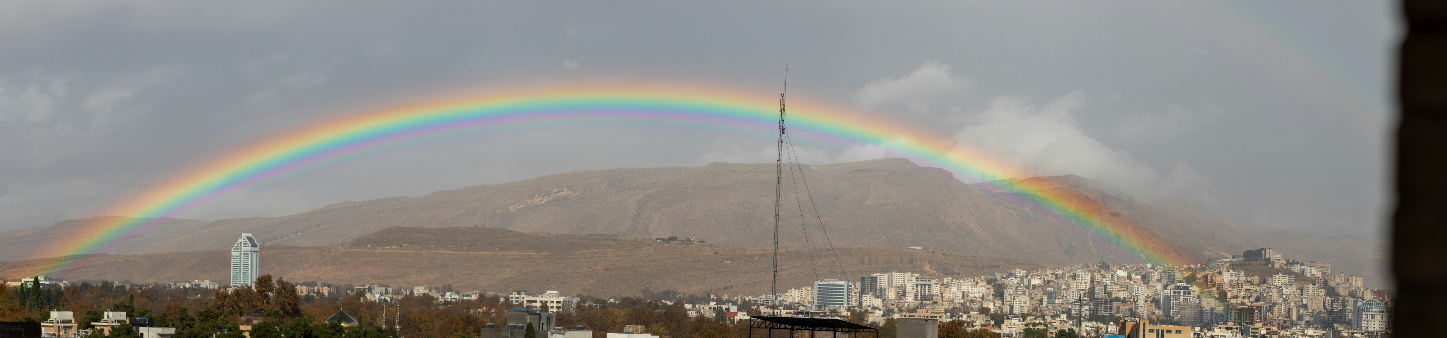 Vibrant rainbow arches over city buildings against a mountainous backdrop with overcast skies.