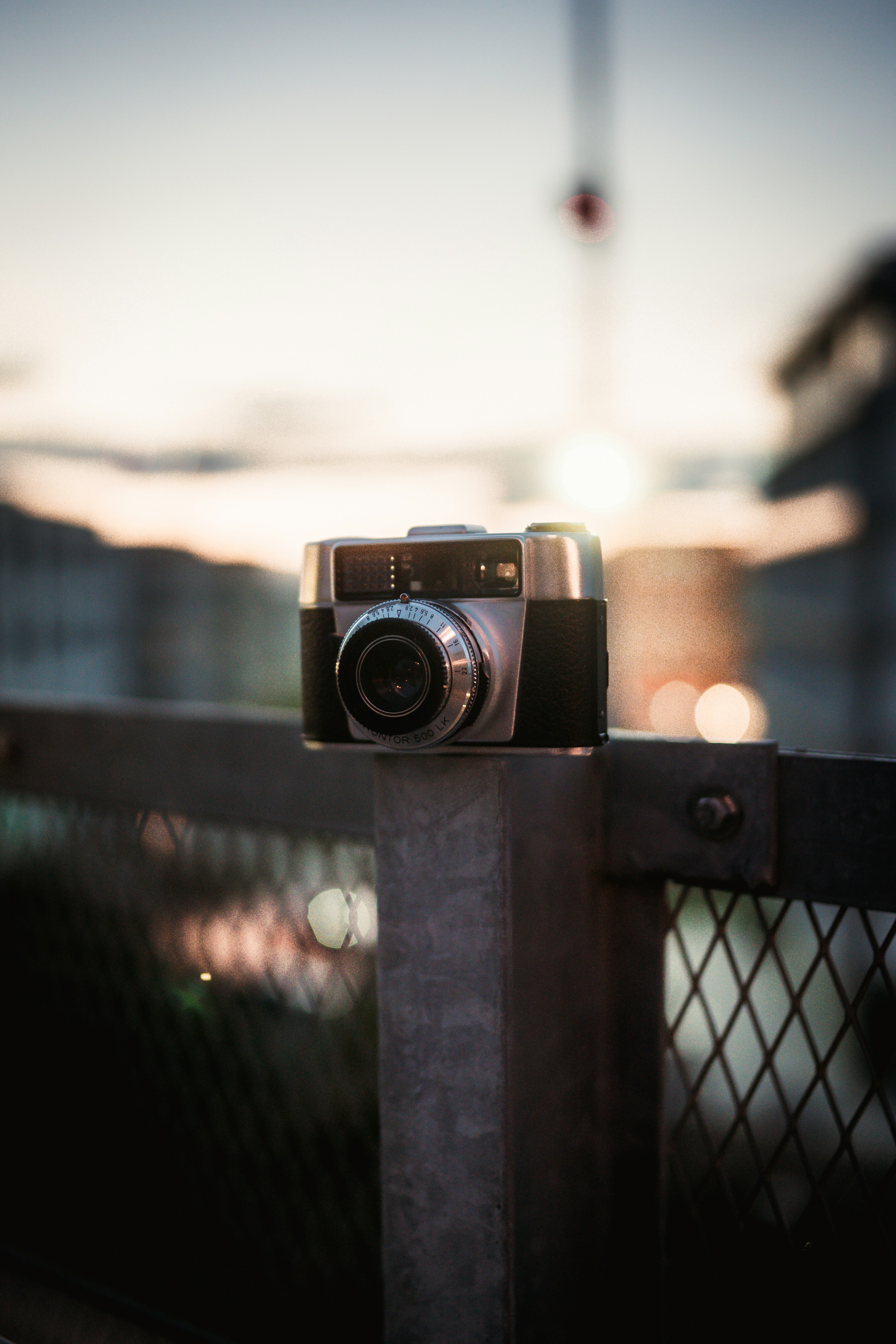 Classic camera resting on a metallic railing, with a soft, blurred cityscape backdrop at dusk.