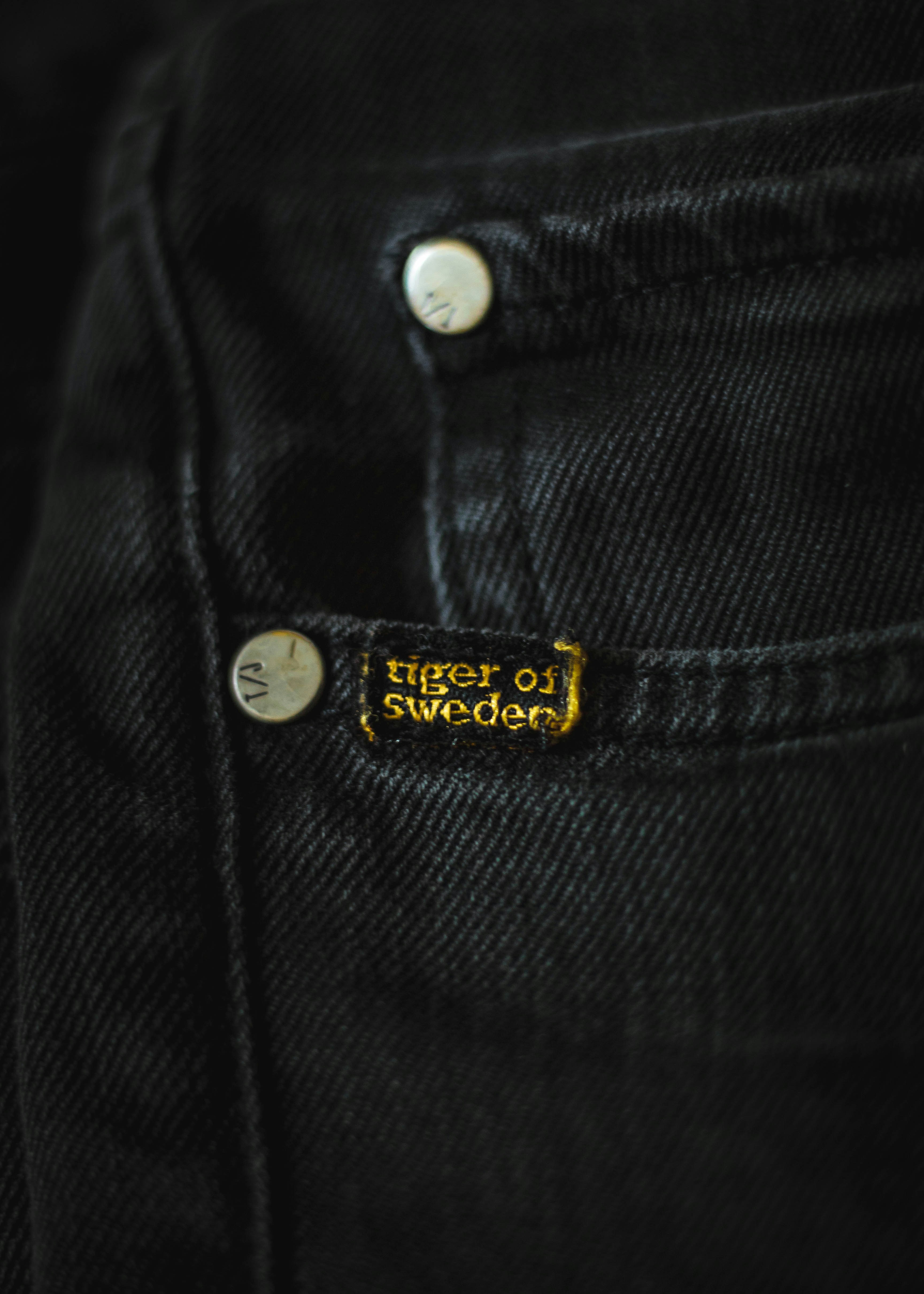 Close-up of a black denim pocket featuring a yellow embroidered label that reads 'Tiger of Sweden.'