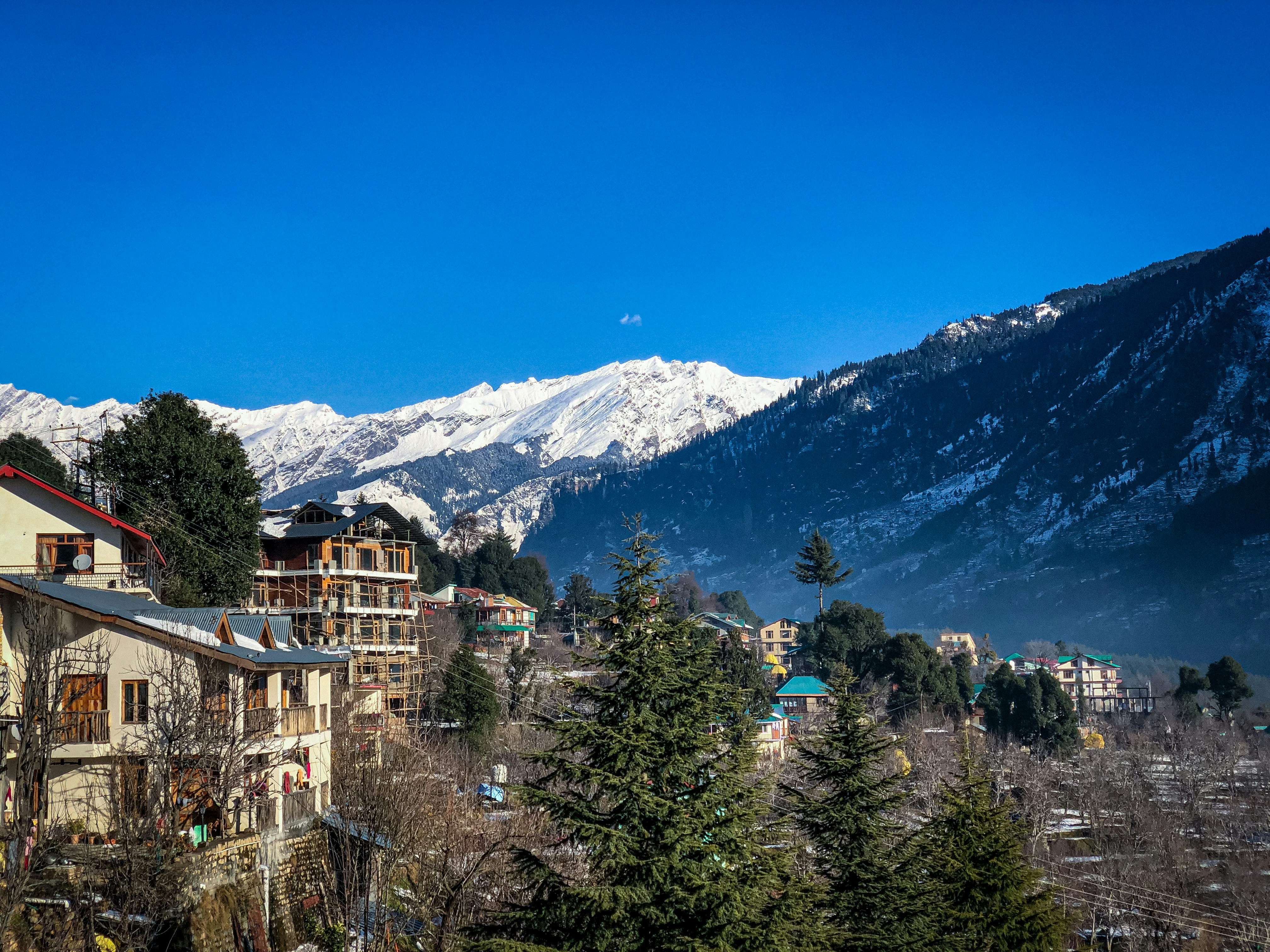 Snow-capped mountains rise majestically above a quaint village nestled in the valley, framed by evergreen trees. The clear blue sky enhances the serene winter landscape.