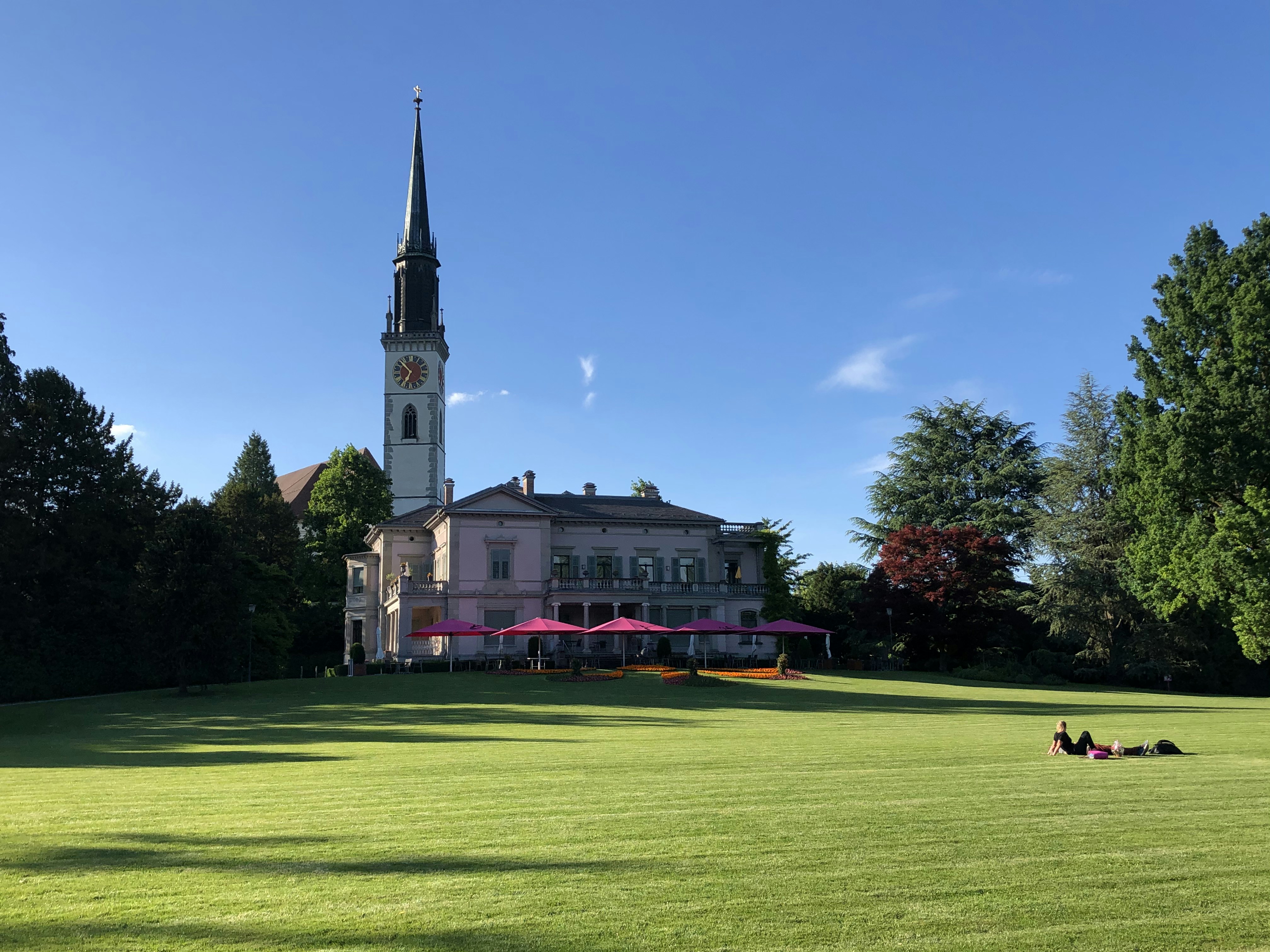 Historic building with a tall spire surrounded by lush greenery under a clear blue sky.