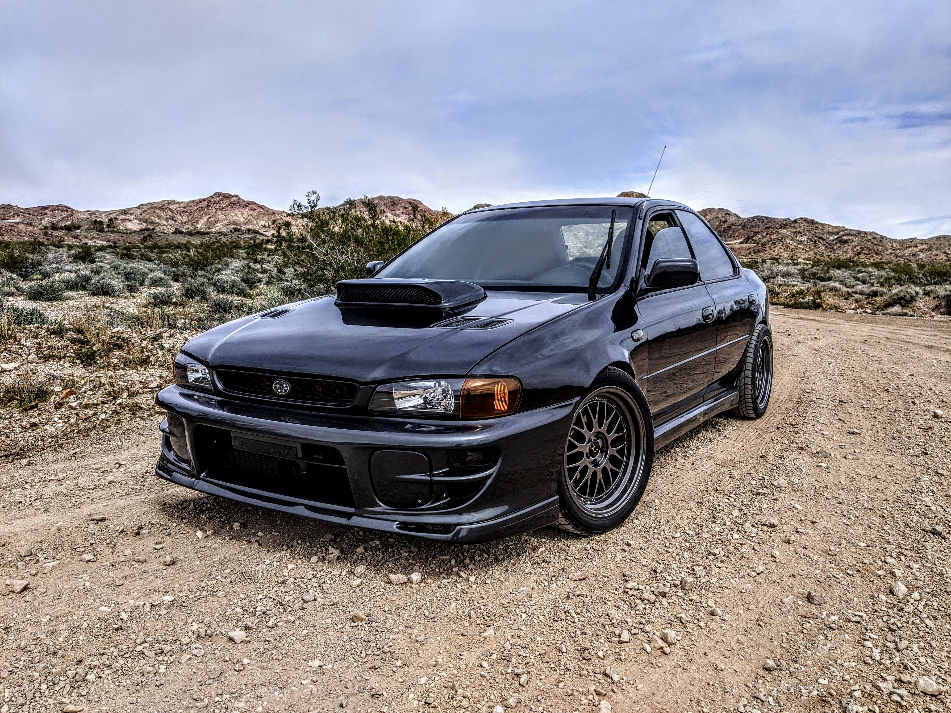 Modified black sports car parked on a rugged dirt road with desert hills in the background.