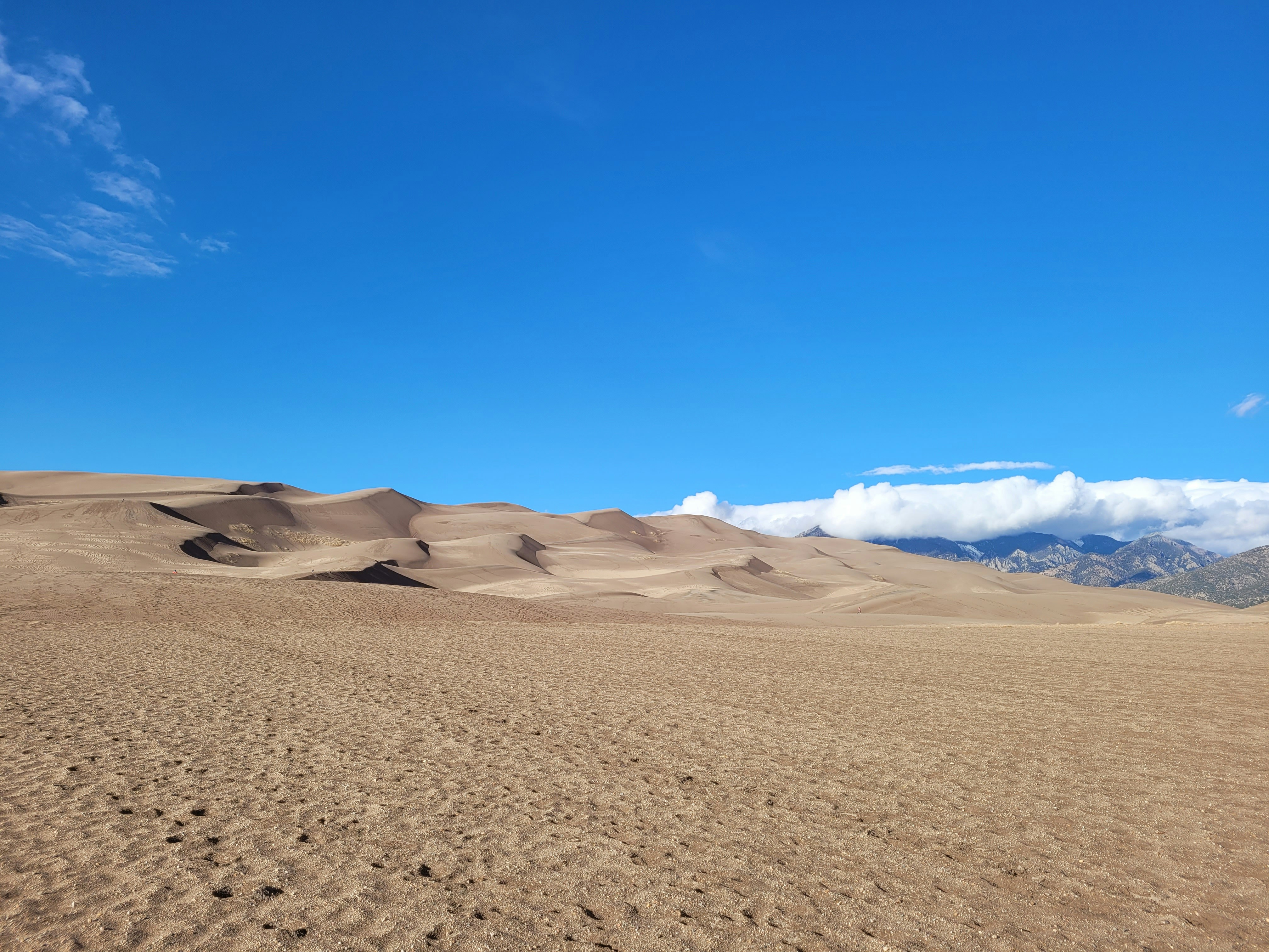 Expansive sandy dunes under a clear blue sky, with distant mountains partially obscured by clouds. The scene embodies tranquility and the vastness of nature.