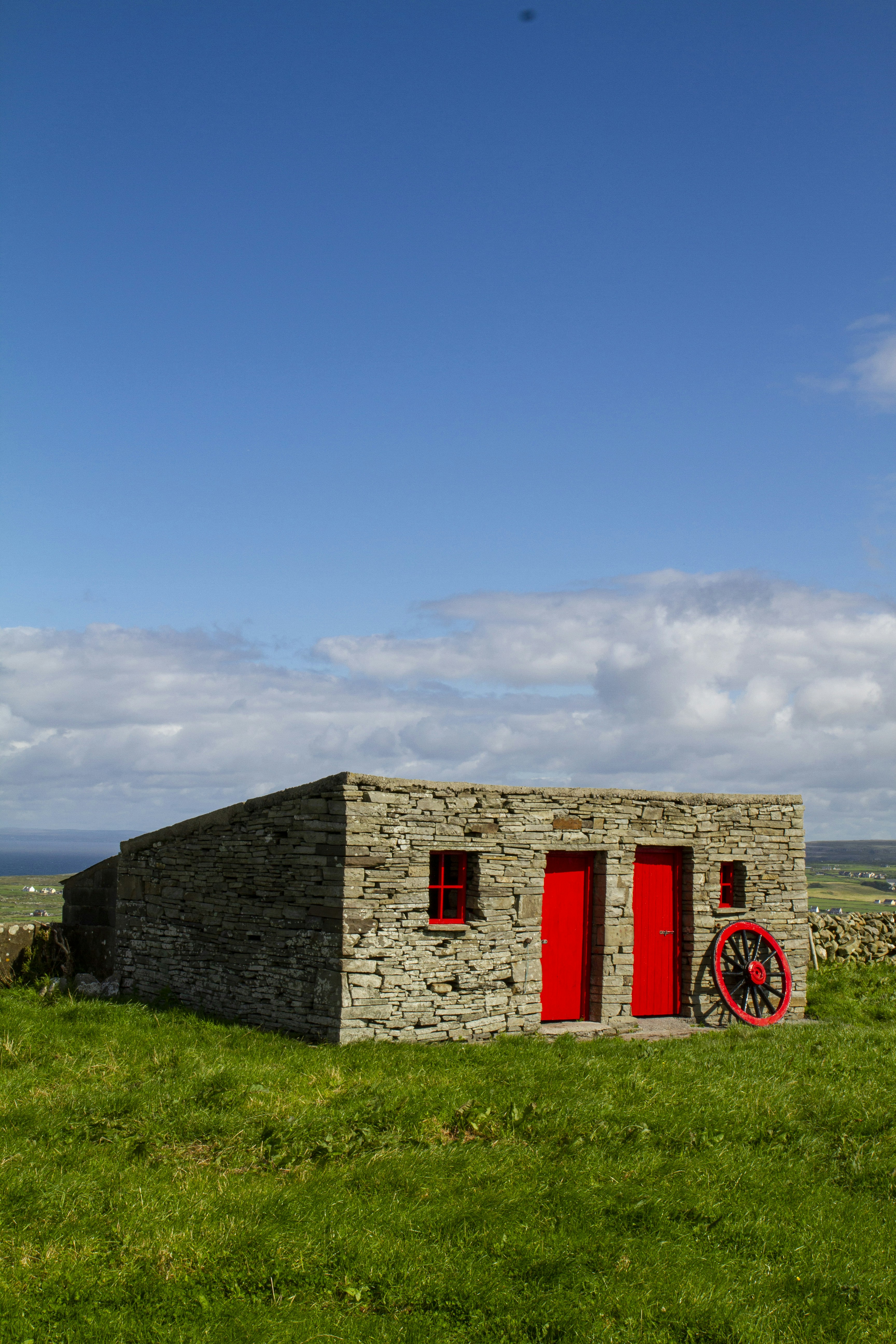A stone cottage with vibrant red doors and windows stands against a bright blue sky, surrounded by lush green grass. The structure features a decorative wheel, adding character to the scene.