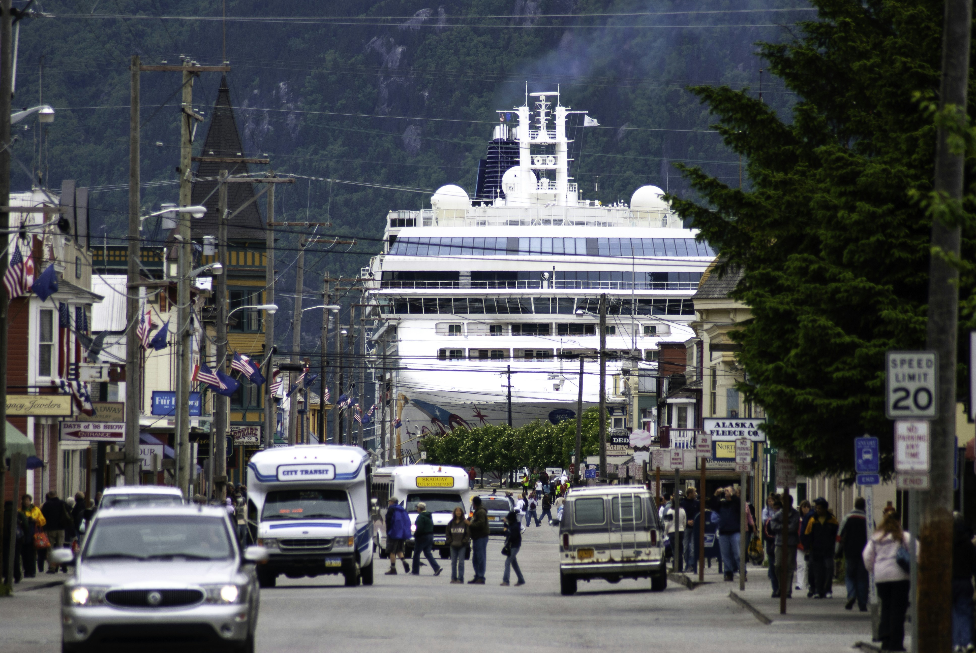 A bustling street scene with pedestrians and vehicles, framed by a majestic cruise ship docked in the background, showcasing the vibrant life of a coastal town.