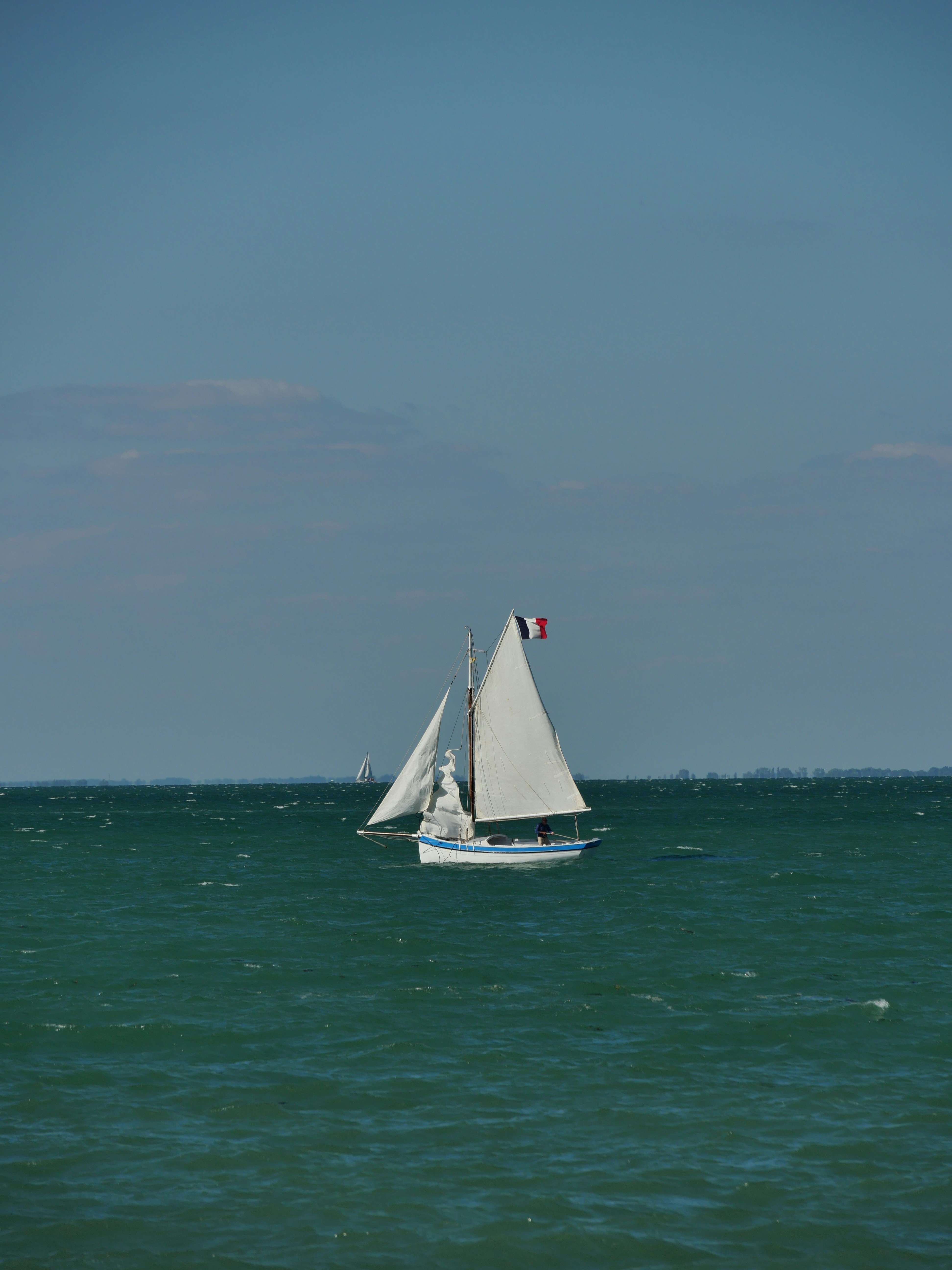 Sailboat gliding across turquoise waters, with white sails billowing under a clear blue sky. French flag proudly displayed at the top.
