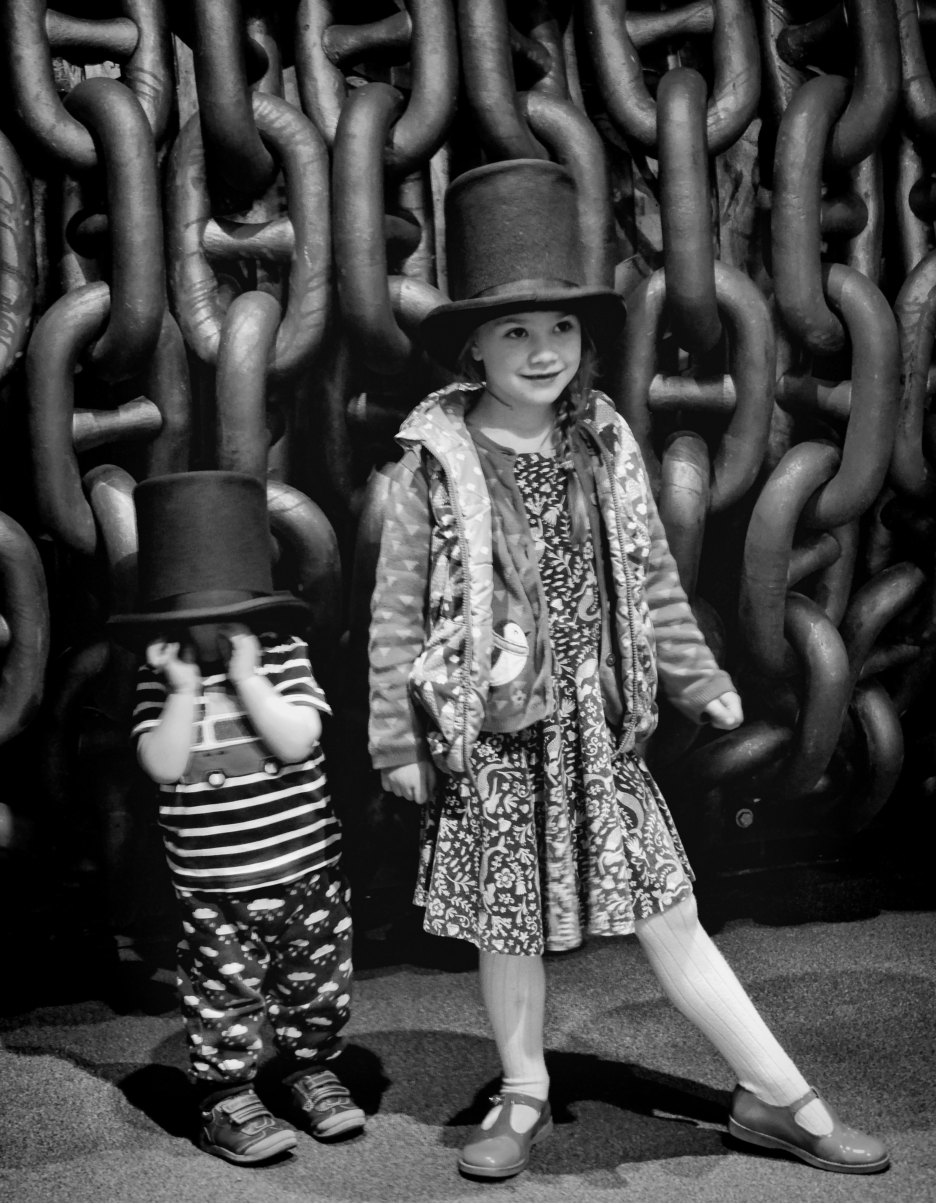 Two children in oversized top hats pose playfully against a backdrop of large chains, showcasing a blend of innocence and imagination.