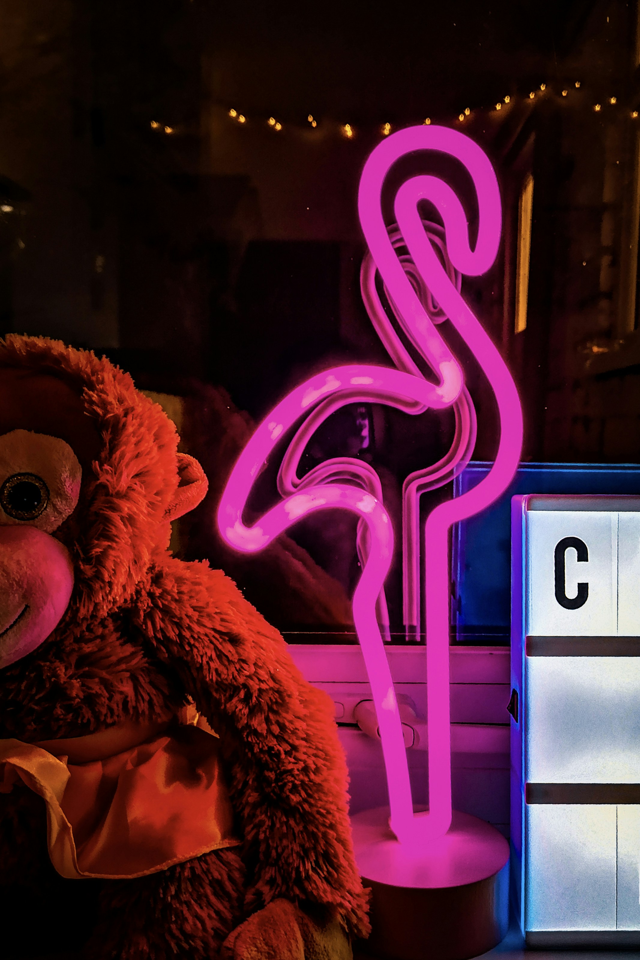 A vibrant neon flamingo lamp casts a pink glow beside a plush monkey, creating a cozy ambiance in a dimly lit room.