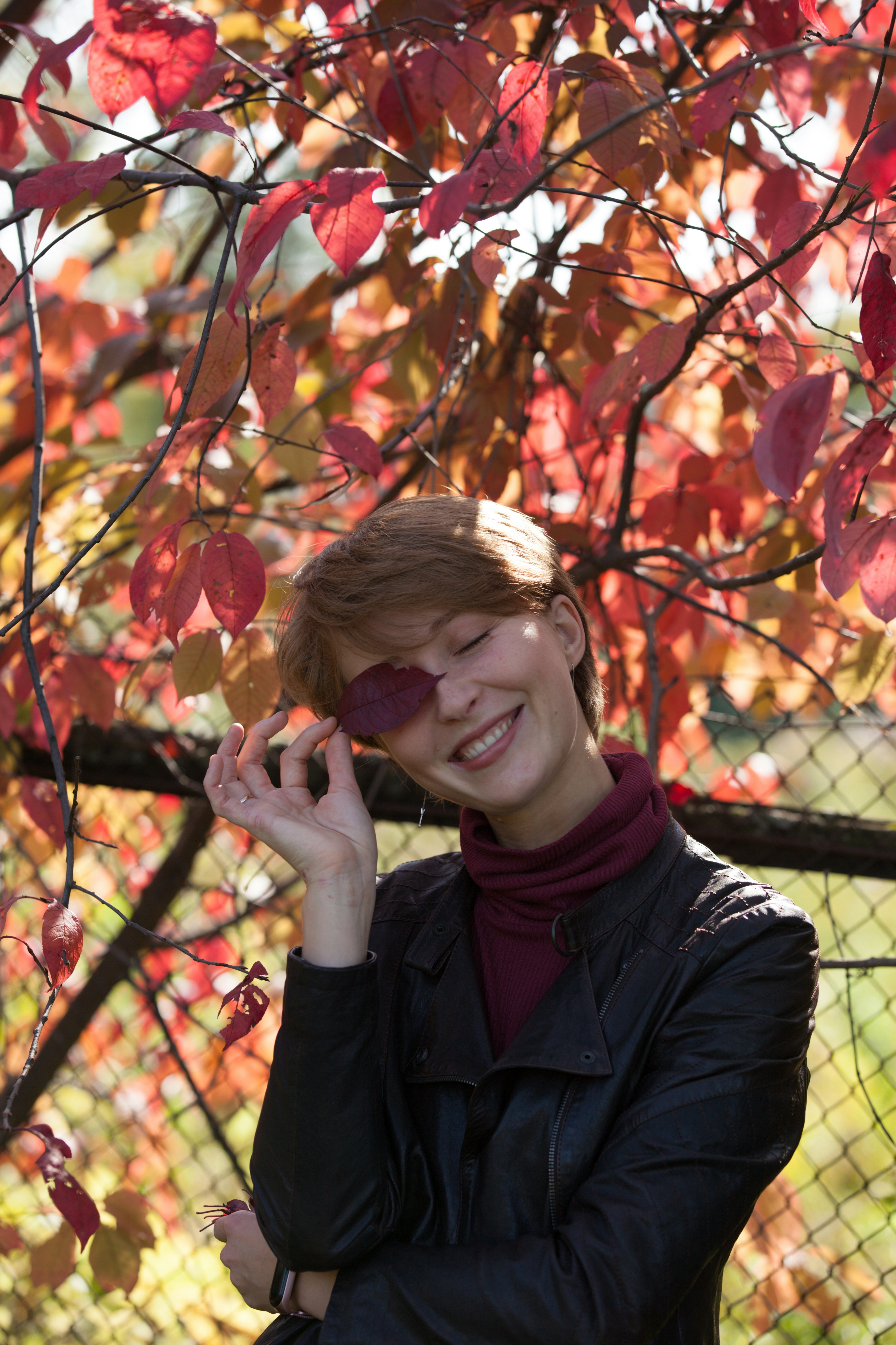 Young woman playfully holding a leaf over her eye, surrounded by vibrant autumn foliage. The scene captures the joy of the season.
