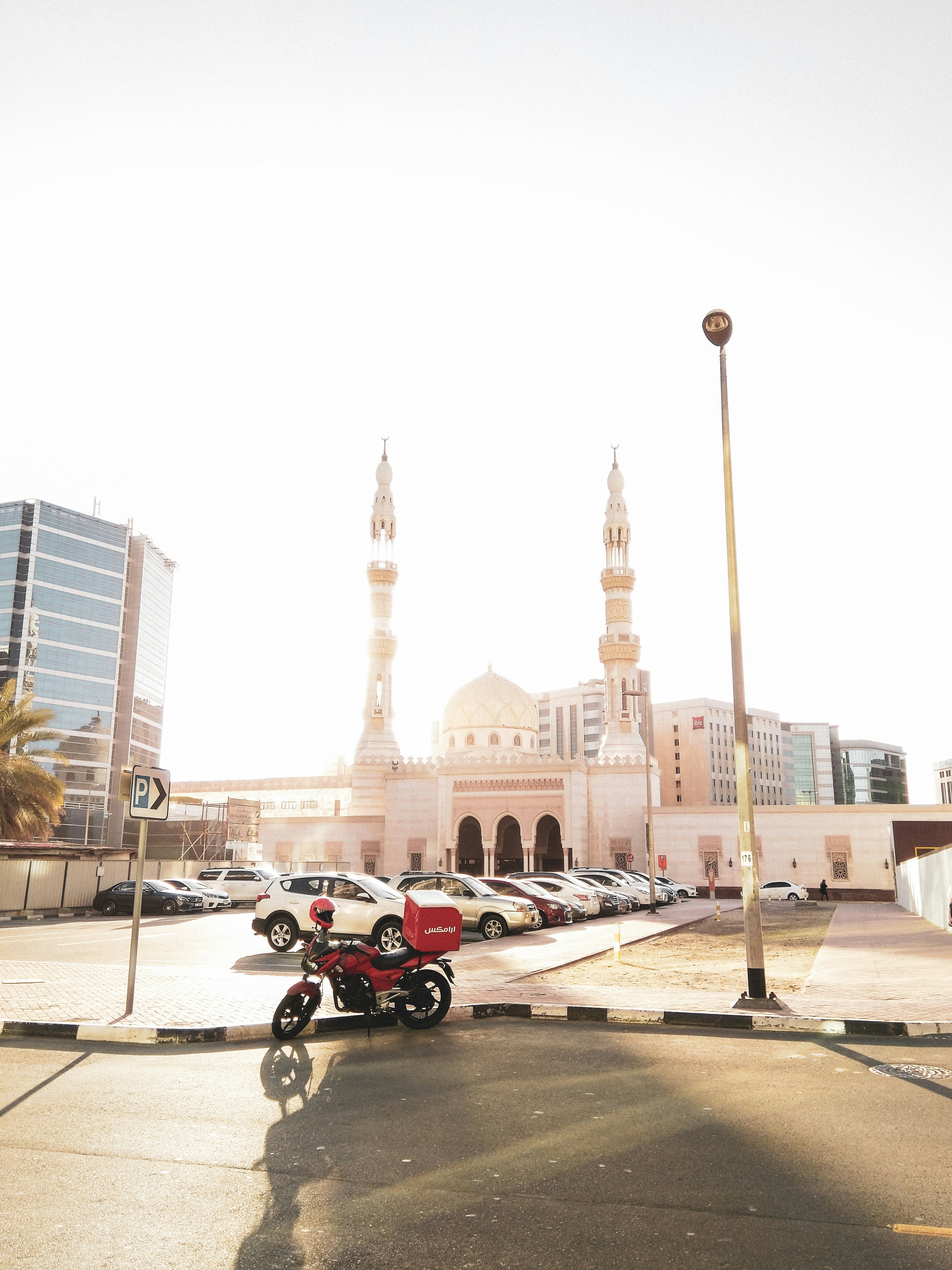 A delivery motorcycle parked in front of a grand mosque, surrounded by modern buildings and cars. The scene captures the blend of urban life and cultural heritage.