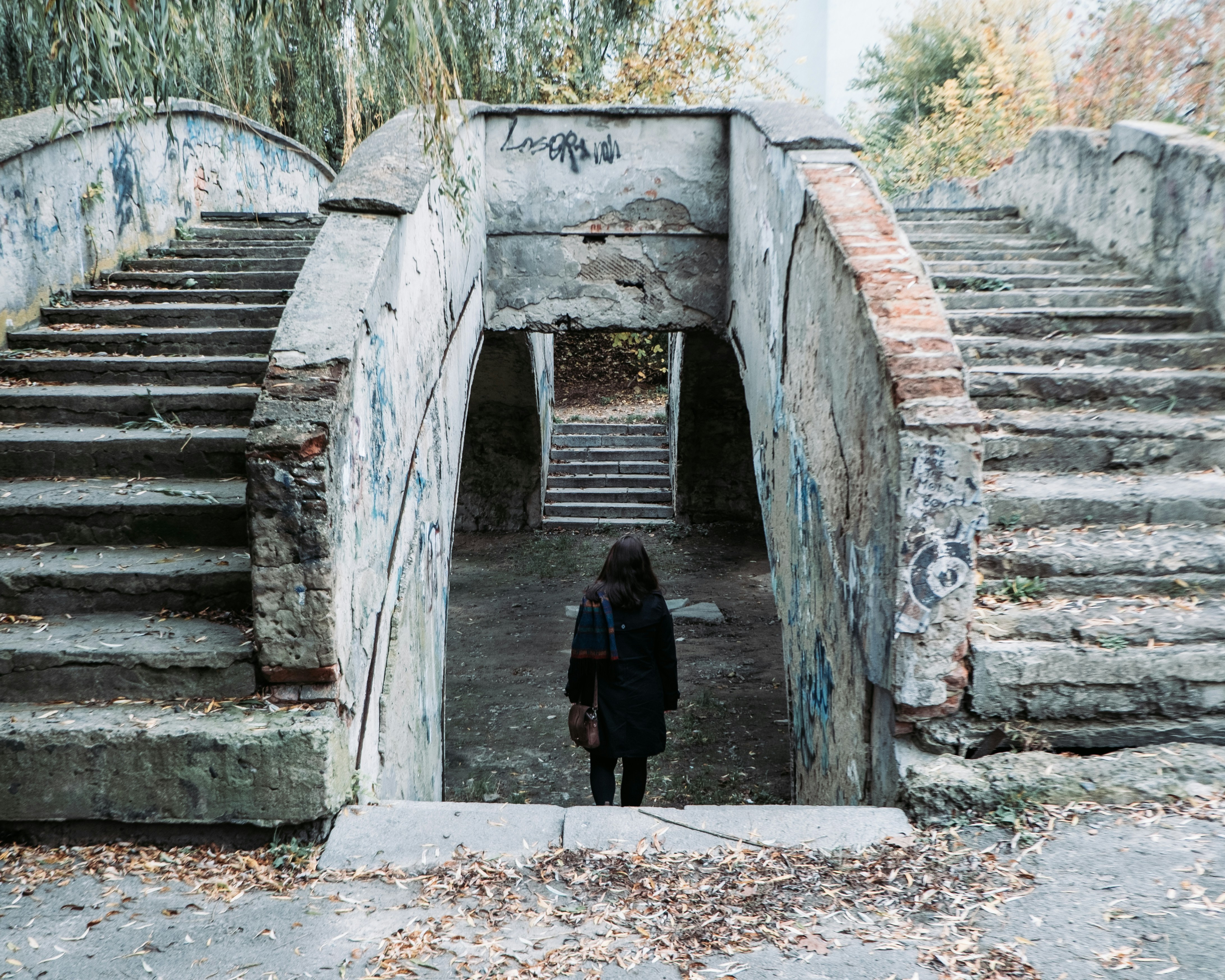 A solitary figure stands at the intersection of two weathered staircases in a graffiti-covered underpass, surrounded by autumn foliage.