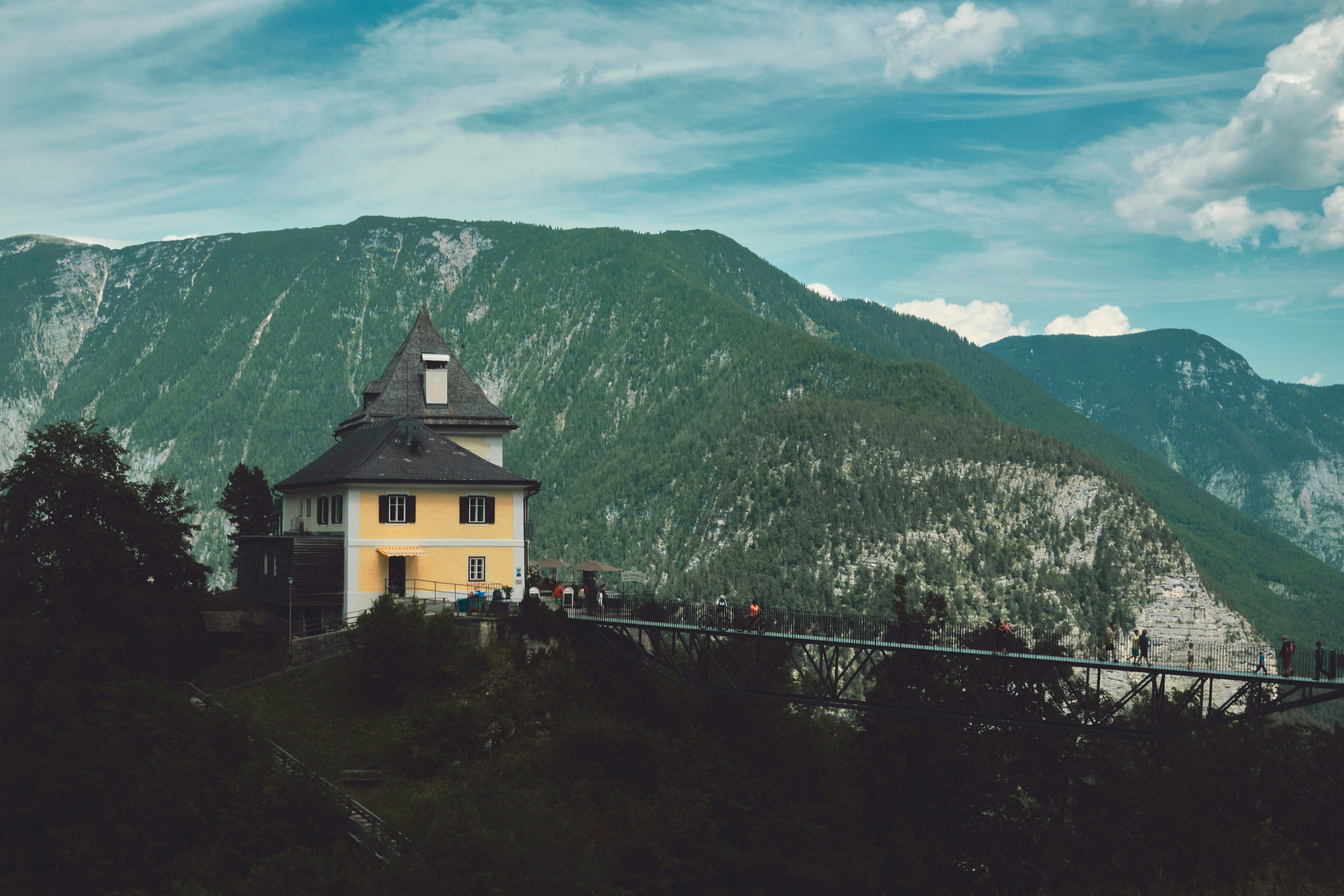 A charming yellow building perched on a ridge, surrounded by lush mountains and a clear blue sky. Visitors stroll along a walkway, taking in the serene landscape.