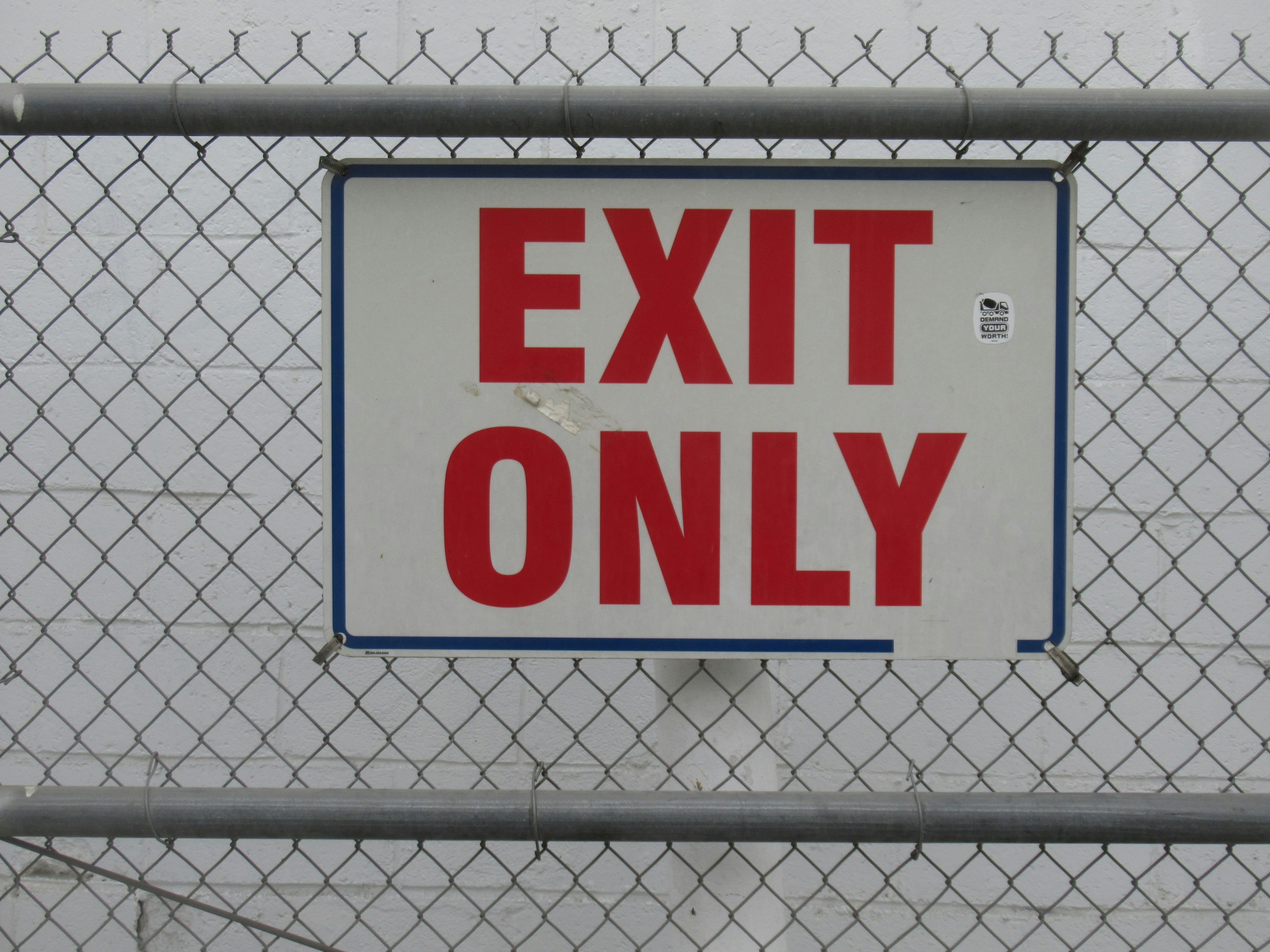 Bold 'EXIT ONLY' sign mounted on a chain-link fence, emphasizing directional guidance in a public space.