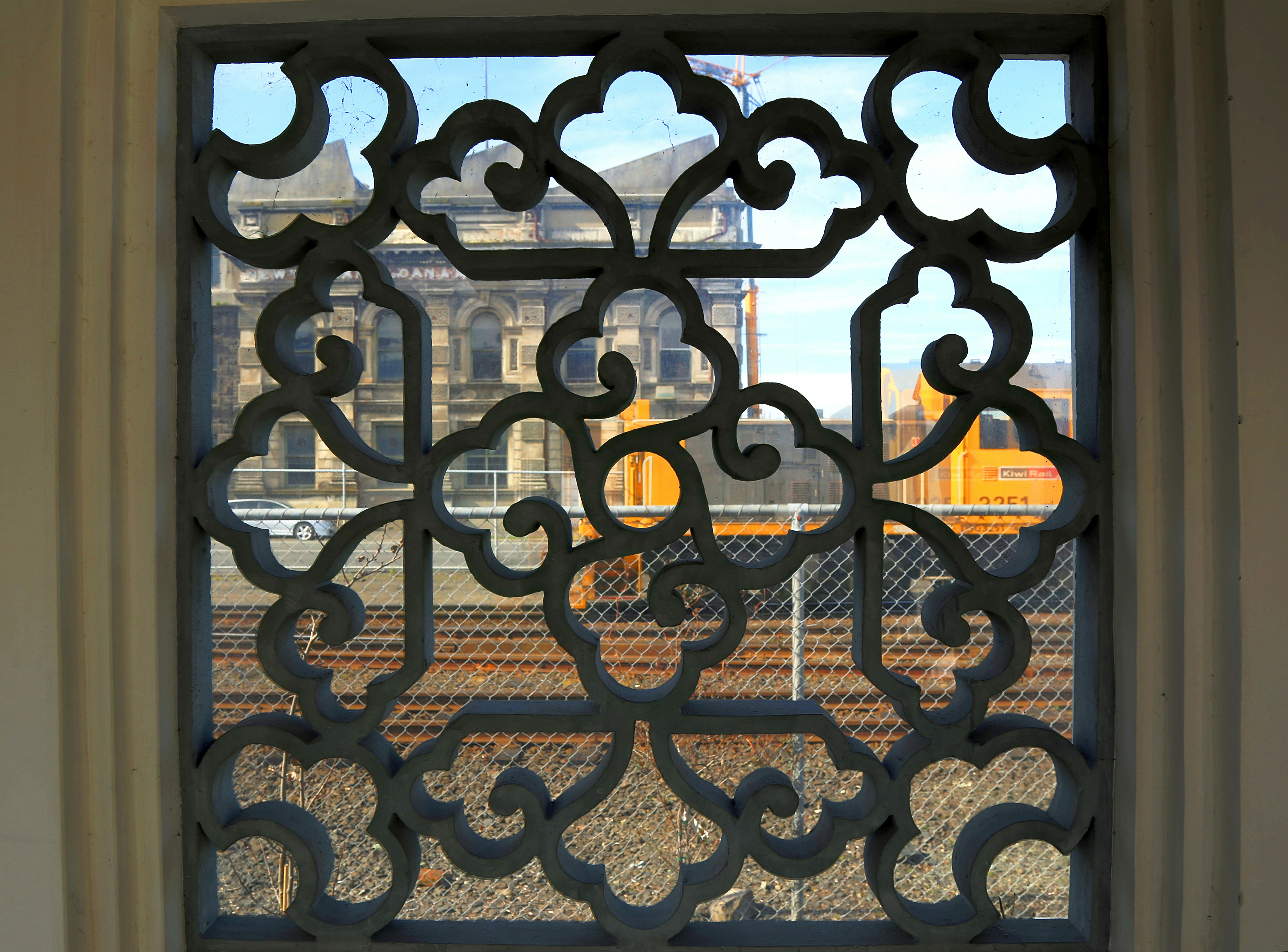 An image of a carved stone window at the Dunedin Chinese Garden. Behind are the railway lines and railway station building. | black metal frame with glass window