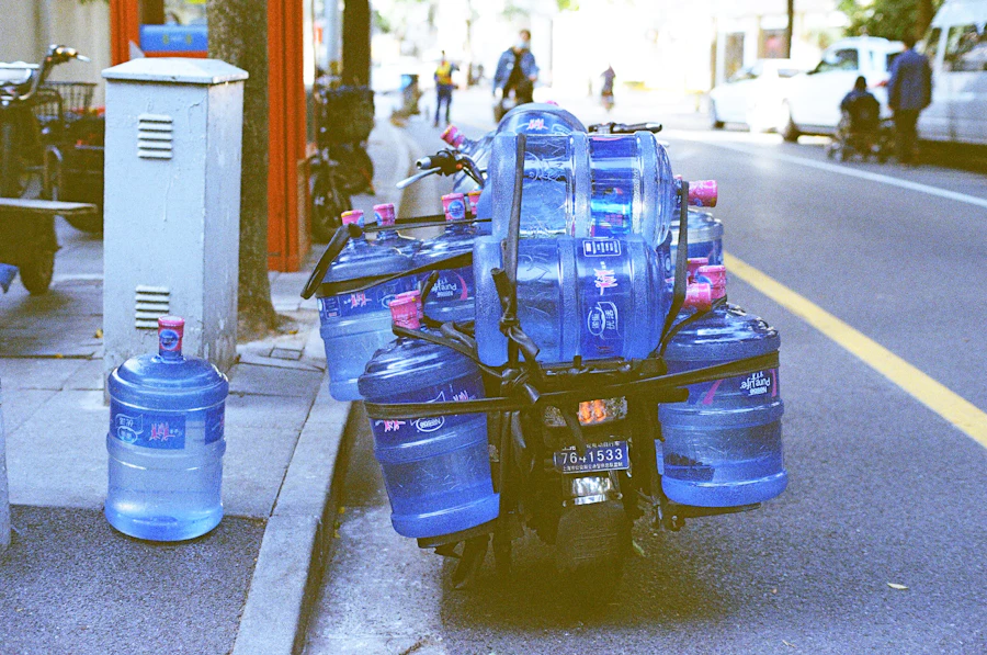 A motorbike parked at the curb is stacked with several large blue water jugs and containers. A city street and blurred pedestrians recede into the bright background.