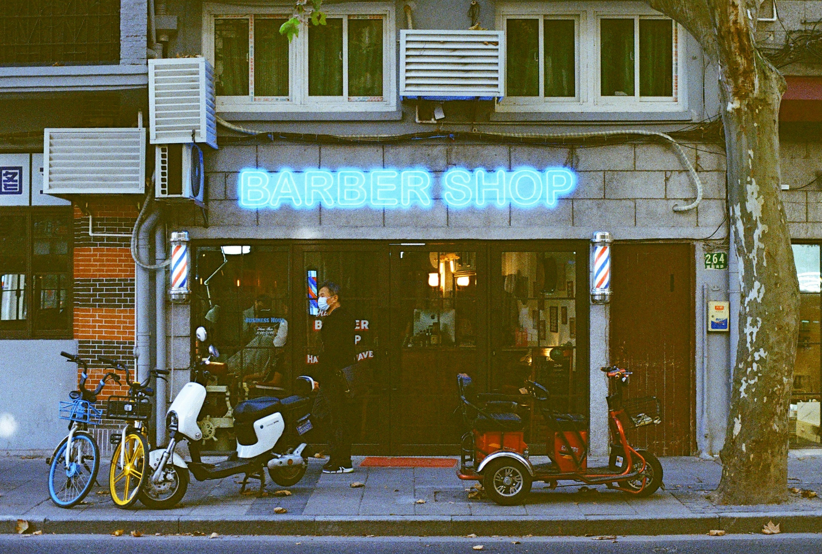 Neon-lit barber shop exterior featuring bicycles and scooters parked outside, showcasing a blend of modern and vintage elements.