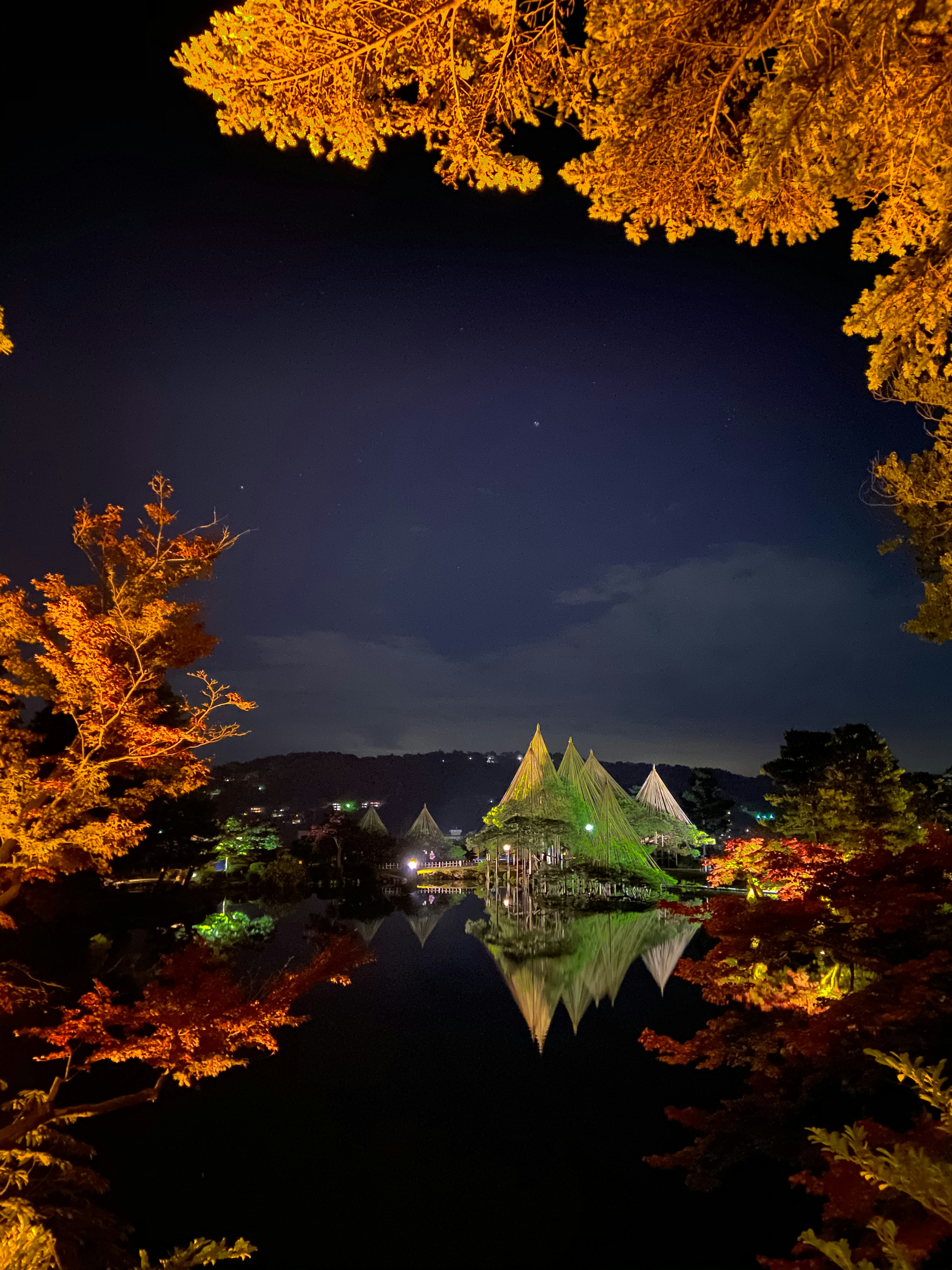 Serene lake at night, framed by vibrant autumn foliage, showcasing traditional structures mirrored in the water's surface.