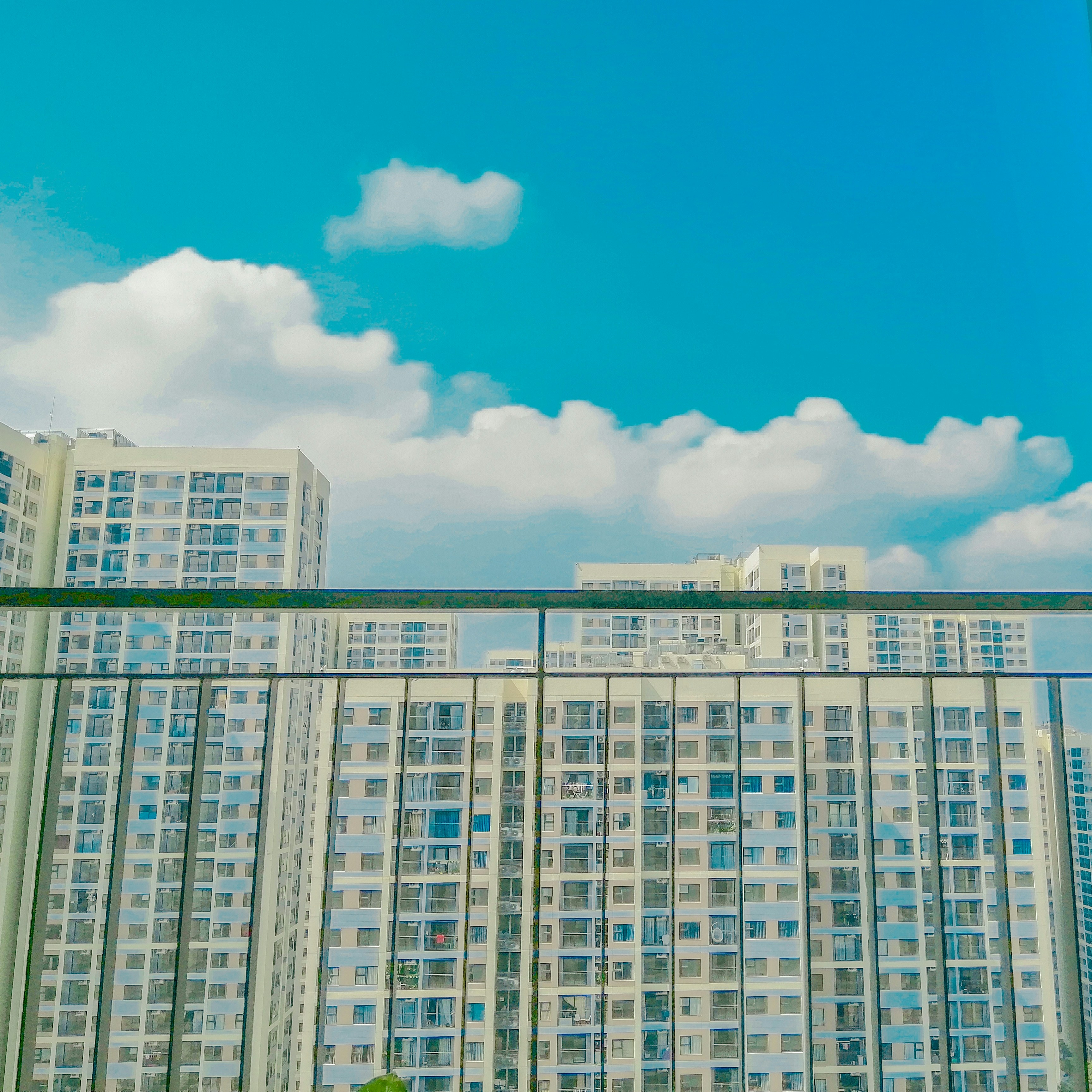 Row of uniform high-rise apartments reflects daylight beneath a bright blue sky, with a metal railing in the foreground.