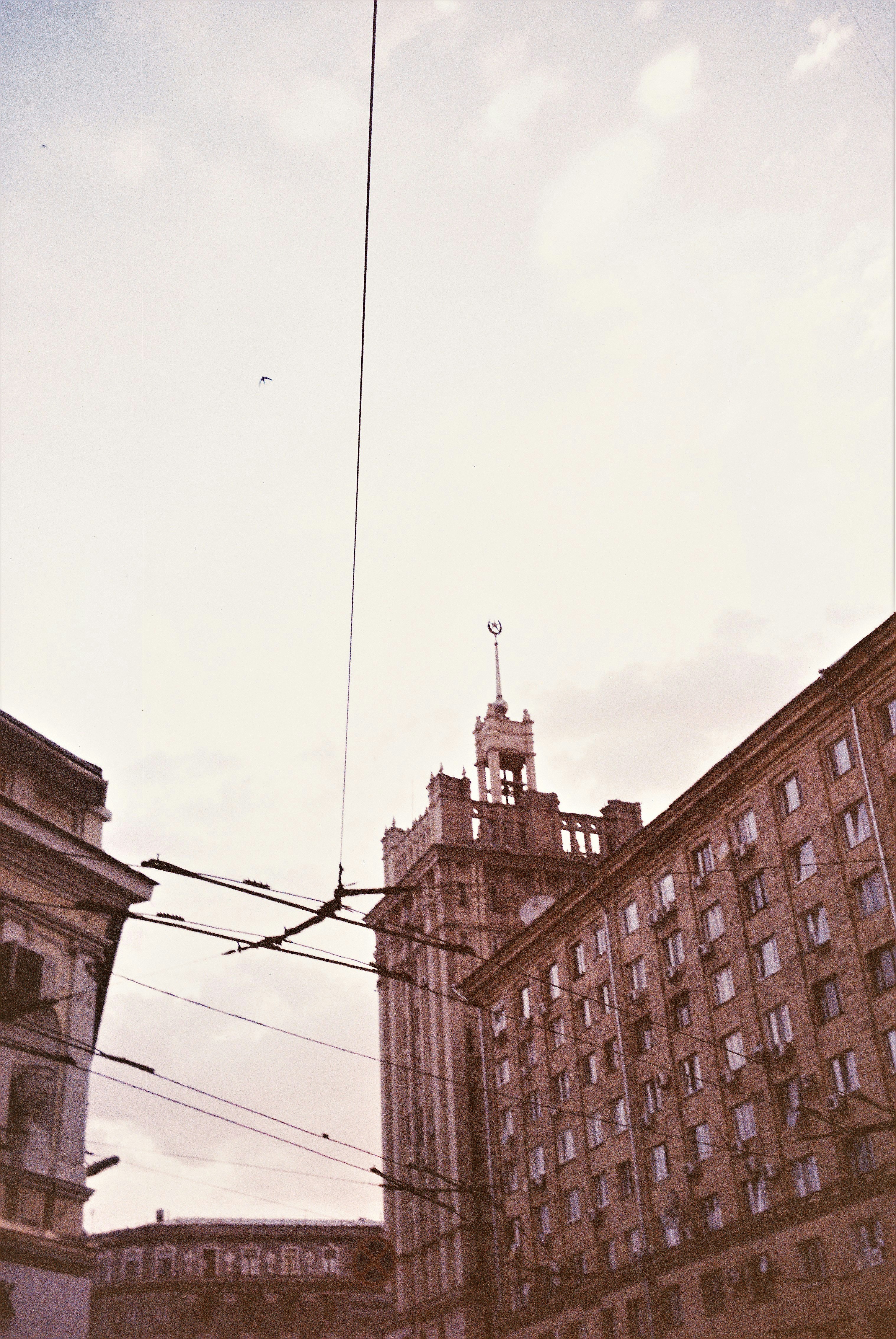 Historic clock tower rising above urban buildings with power lines crisscrossing the scene under a pastel sky.