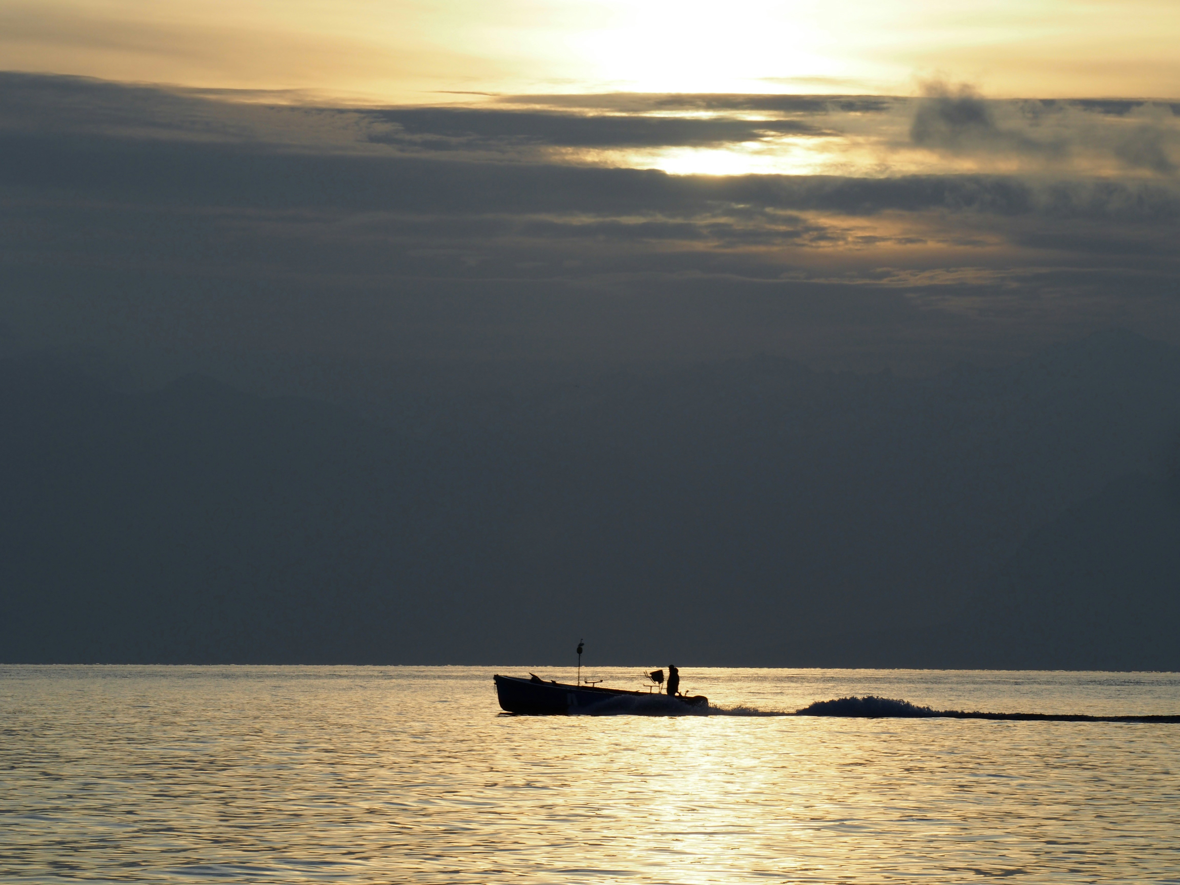 Silhouette of a boat gliding across calm water under a sunlit sky at dusk.