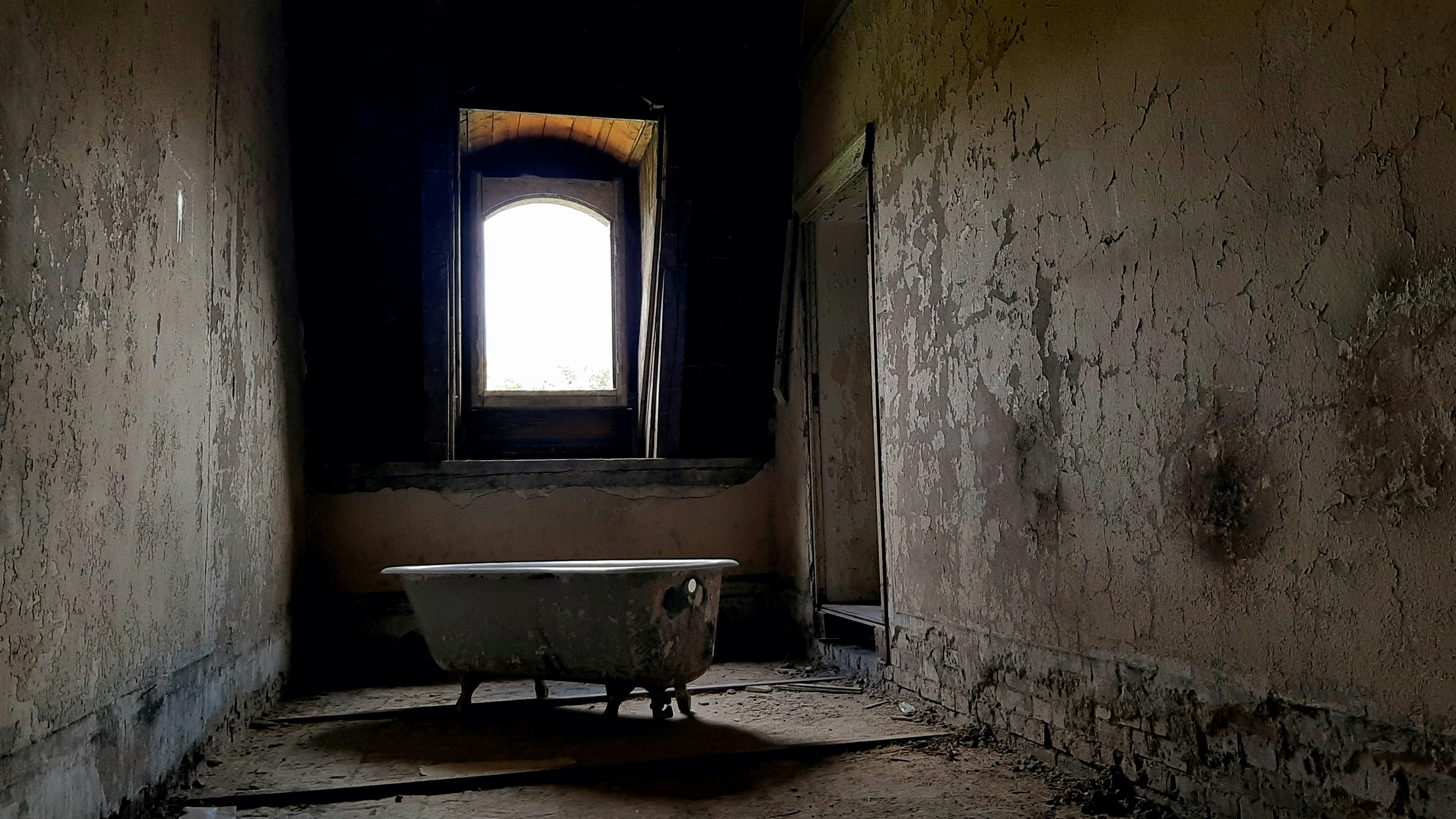 An empty, vintage bathtub sits in the center of a dilapidated room, illuminated by a softly glowing window. The peeling walls and neglected floor evoke a sense of forgotten history.