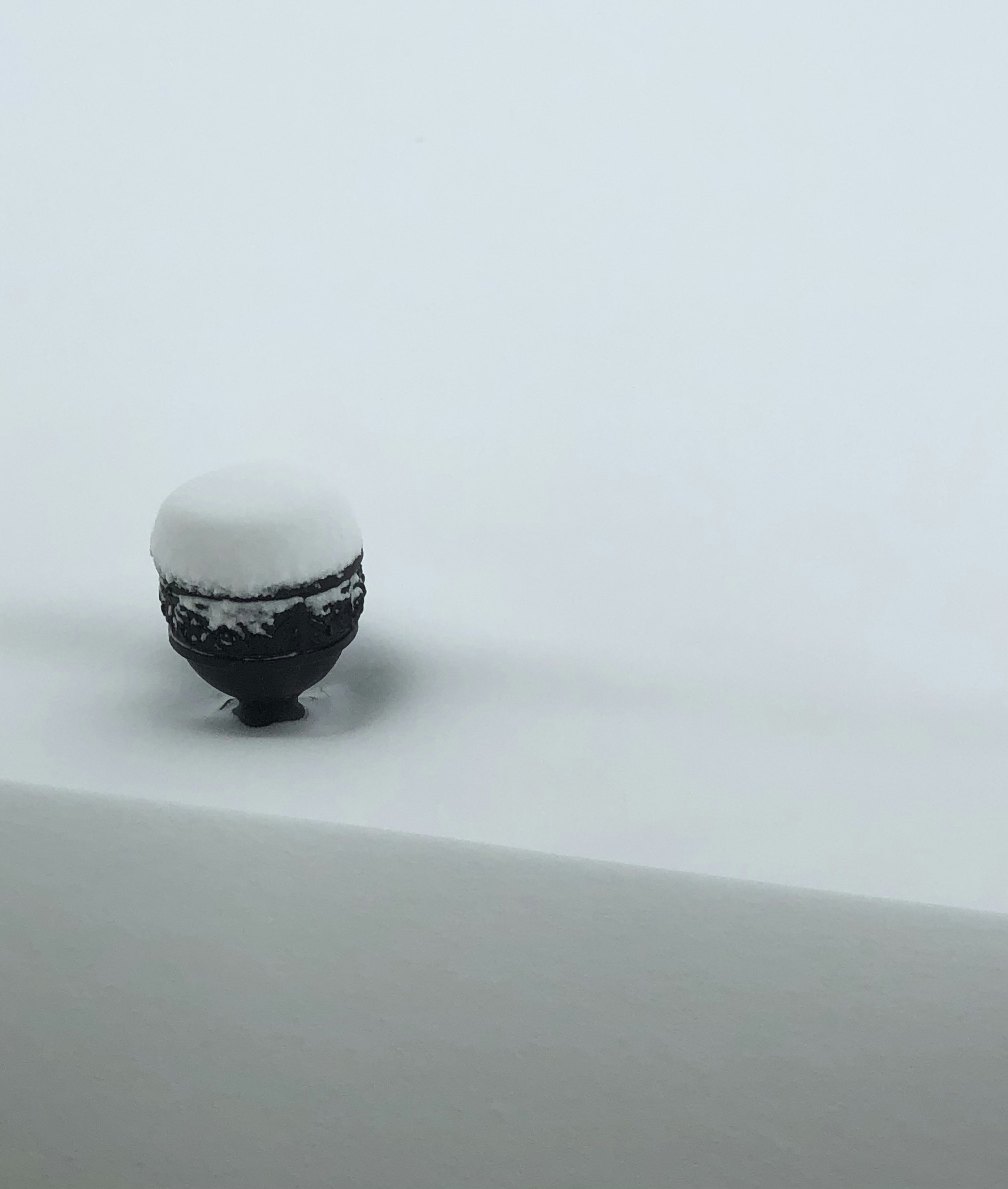 A small, ornate black post is topped with a layer of fresh snow, contrasting against a white background of untouched snow. The scene evokes a sense of stillness and winter's embrace.