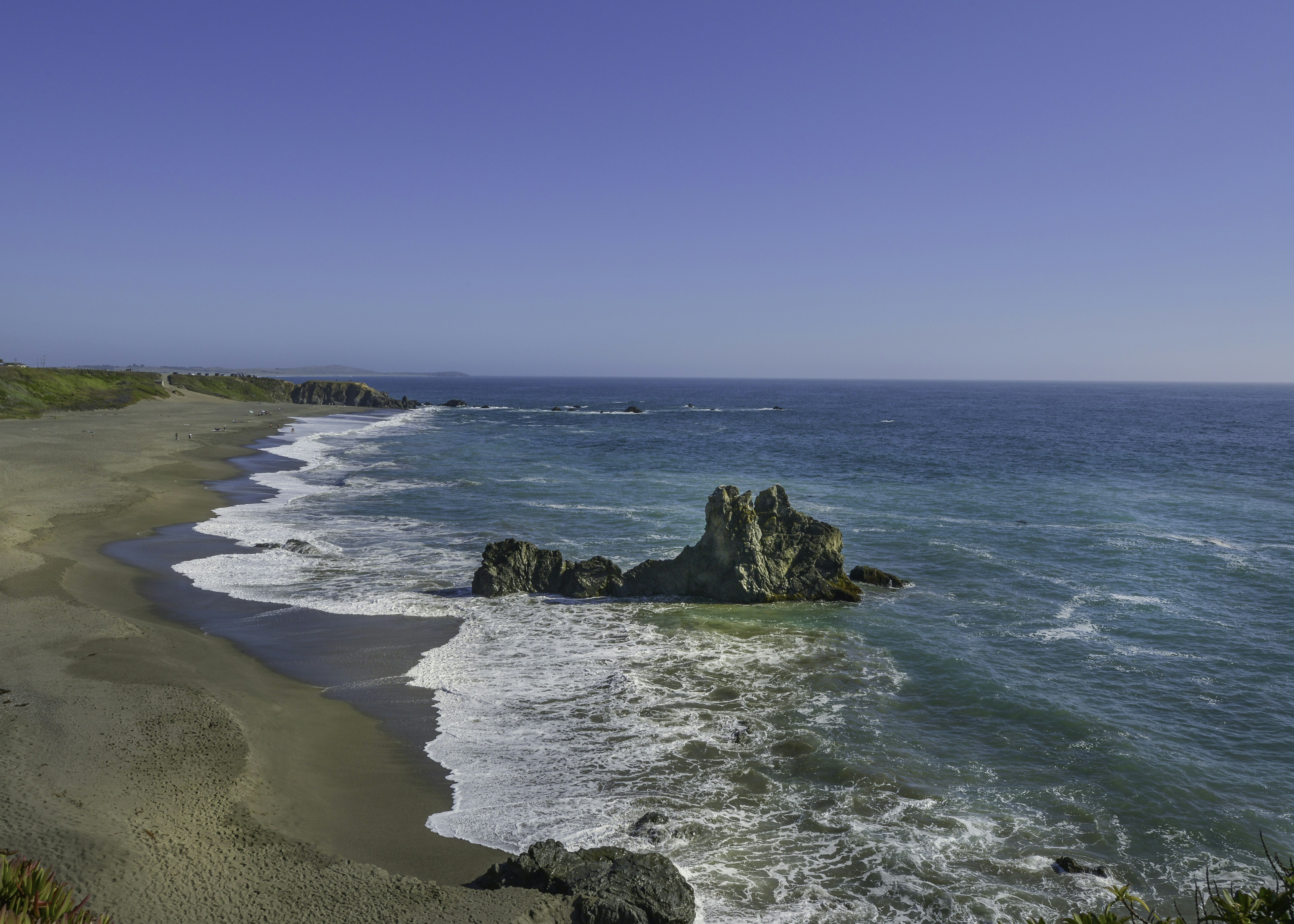 Coastal waves gently lap against a sandy beach with rocky outcrops under a clear blue sky.