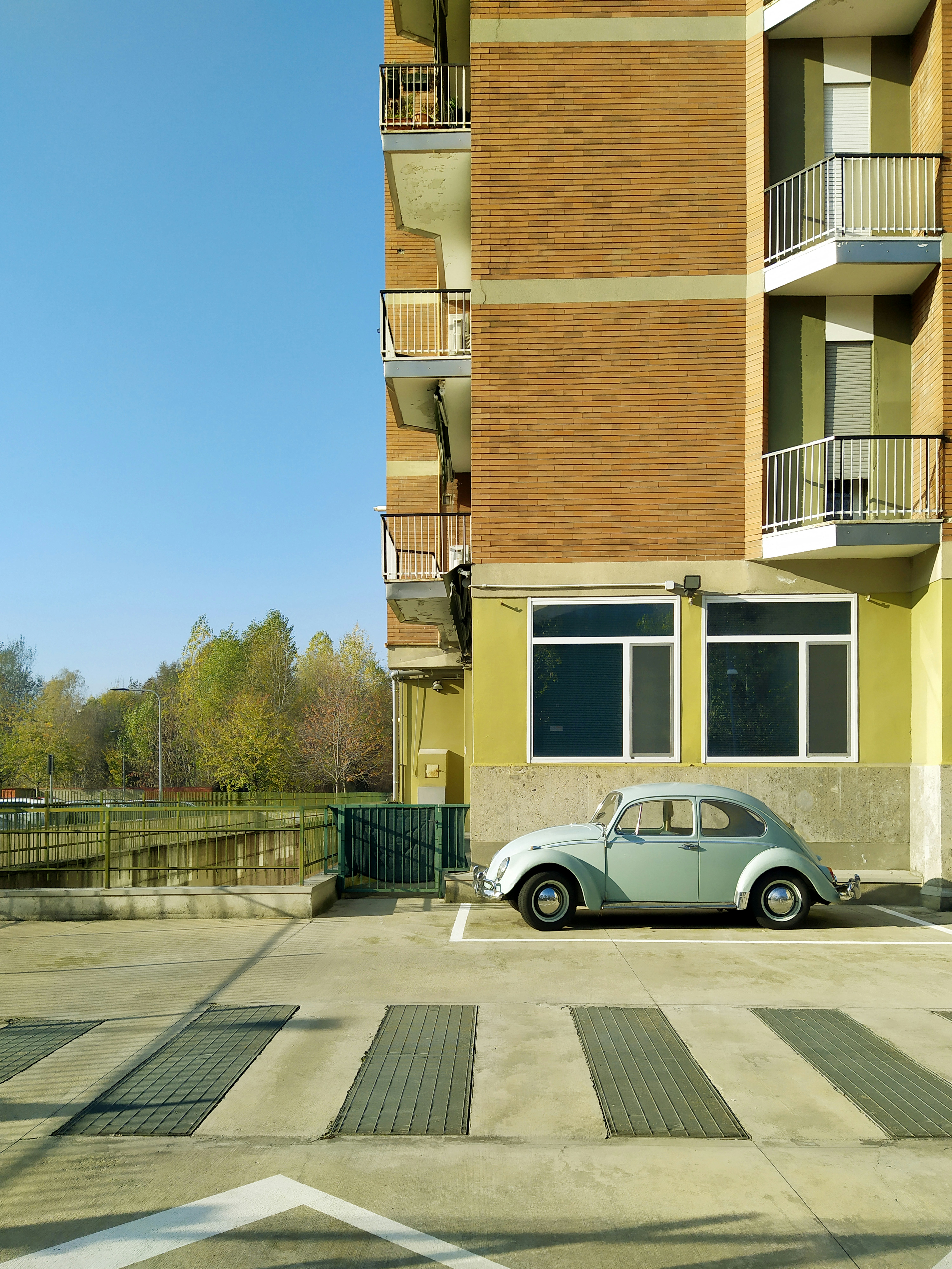 A vintage pale-green Volkswagen Beetle parked beside a sunlit brick apartment block, framed by balconies and a concrete foreground.
