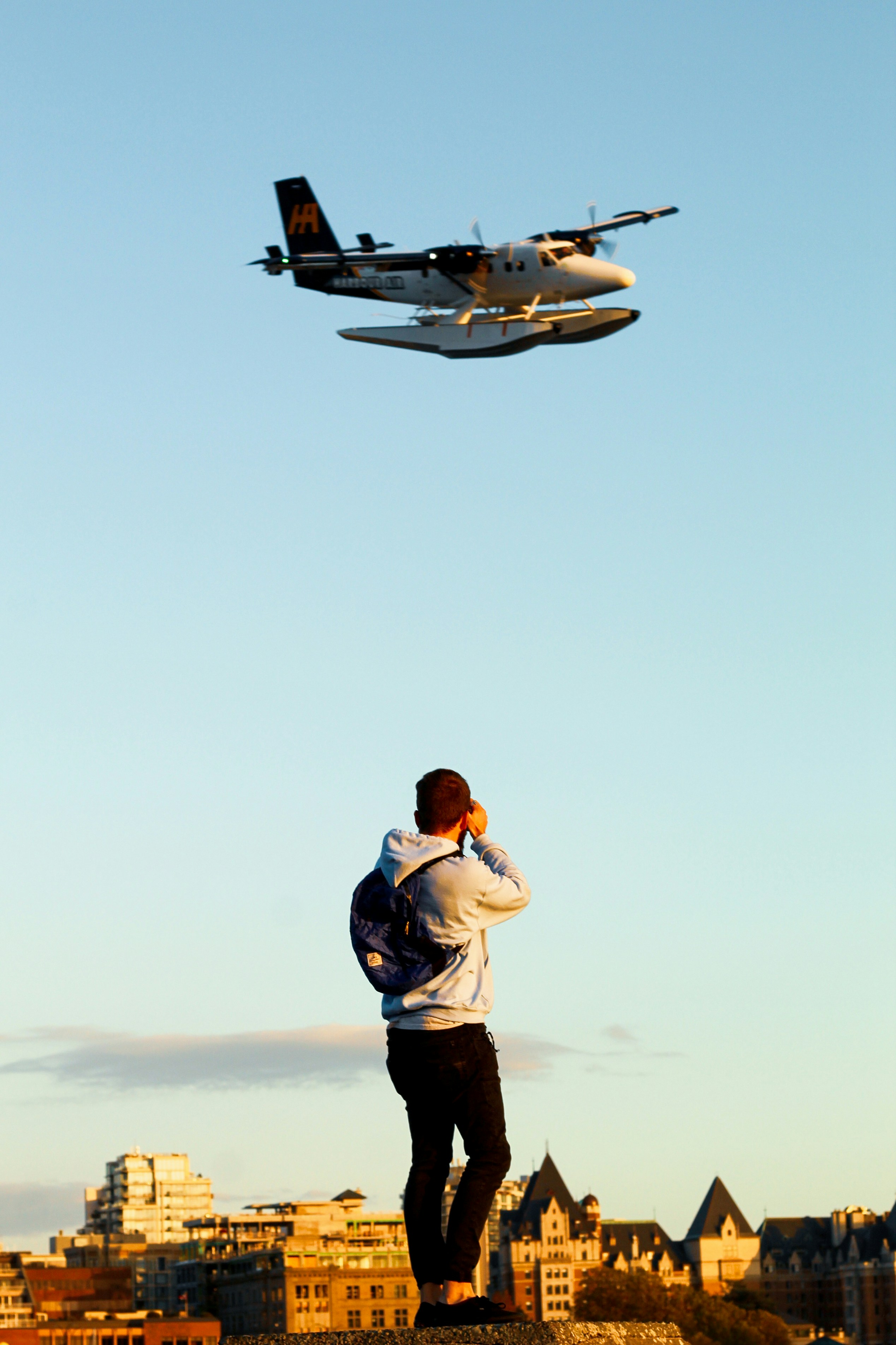 A photographer aims their camera at a seaplane flying overhead against a clear blue sky, with a city skyline in the background.