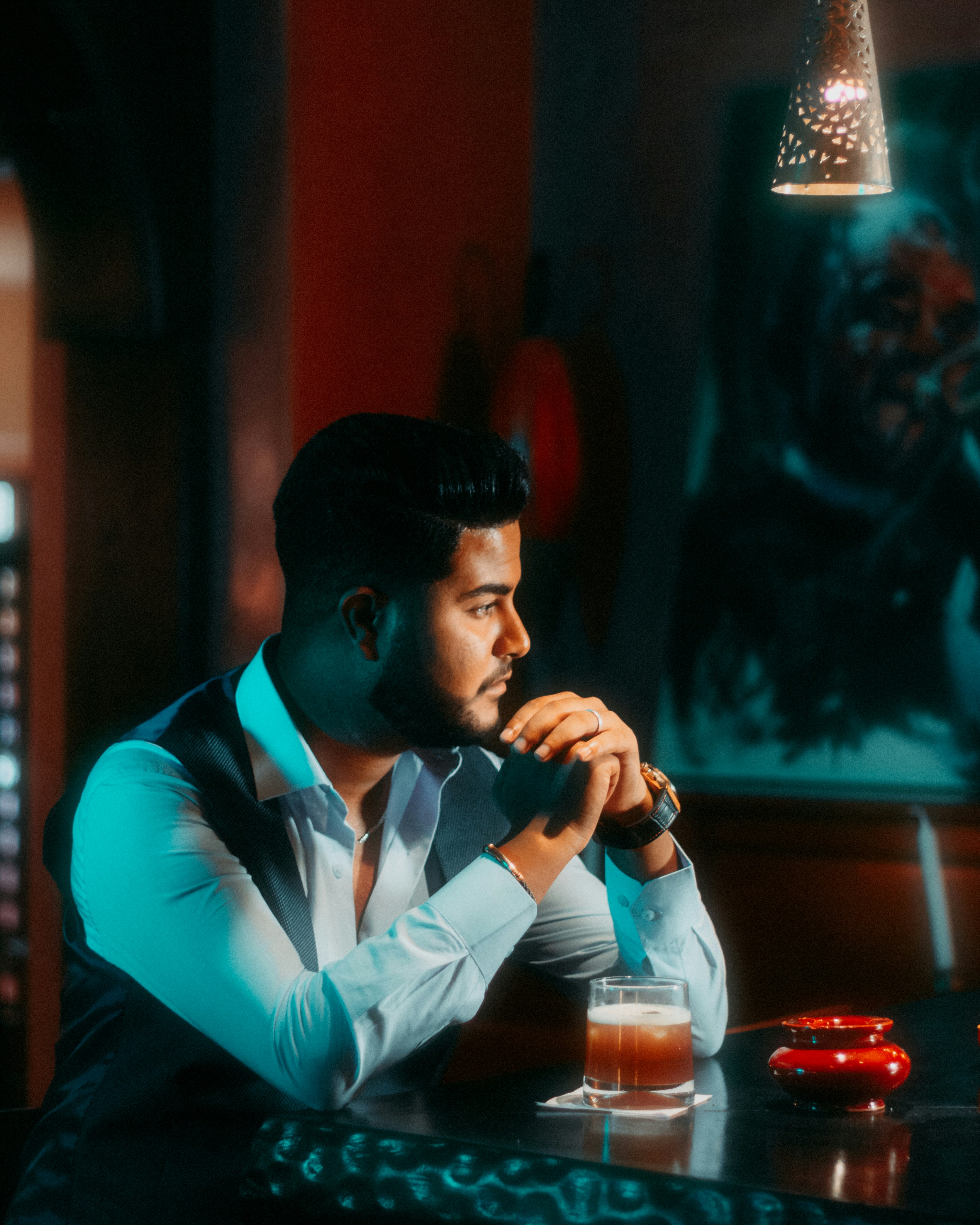 A man sits thoughtfully at a bar, gazing into the distance while holding a drink. The ambient lighting creates a moody atmosphere, enhancing the scene's introspective mood.