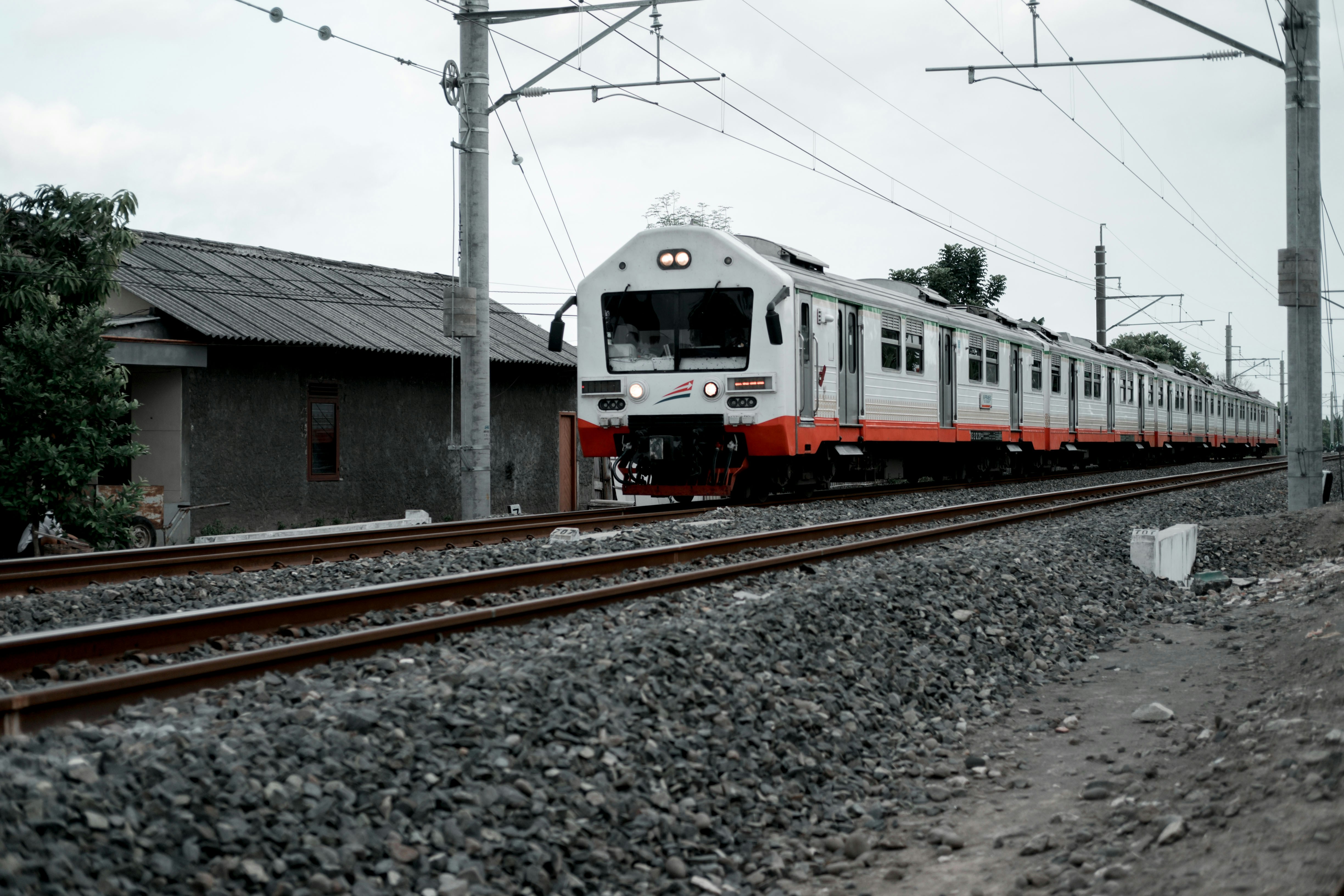 A commuter train approaches a rural station, flanked by traditional architecture and a gravel track. The scene captures the intersection of modern transit and local life.